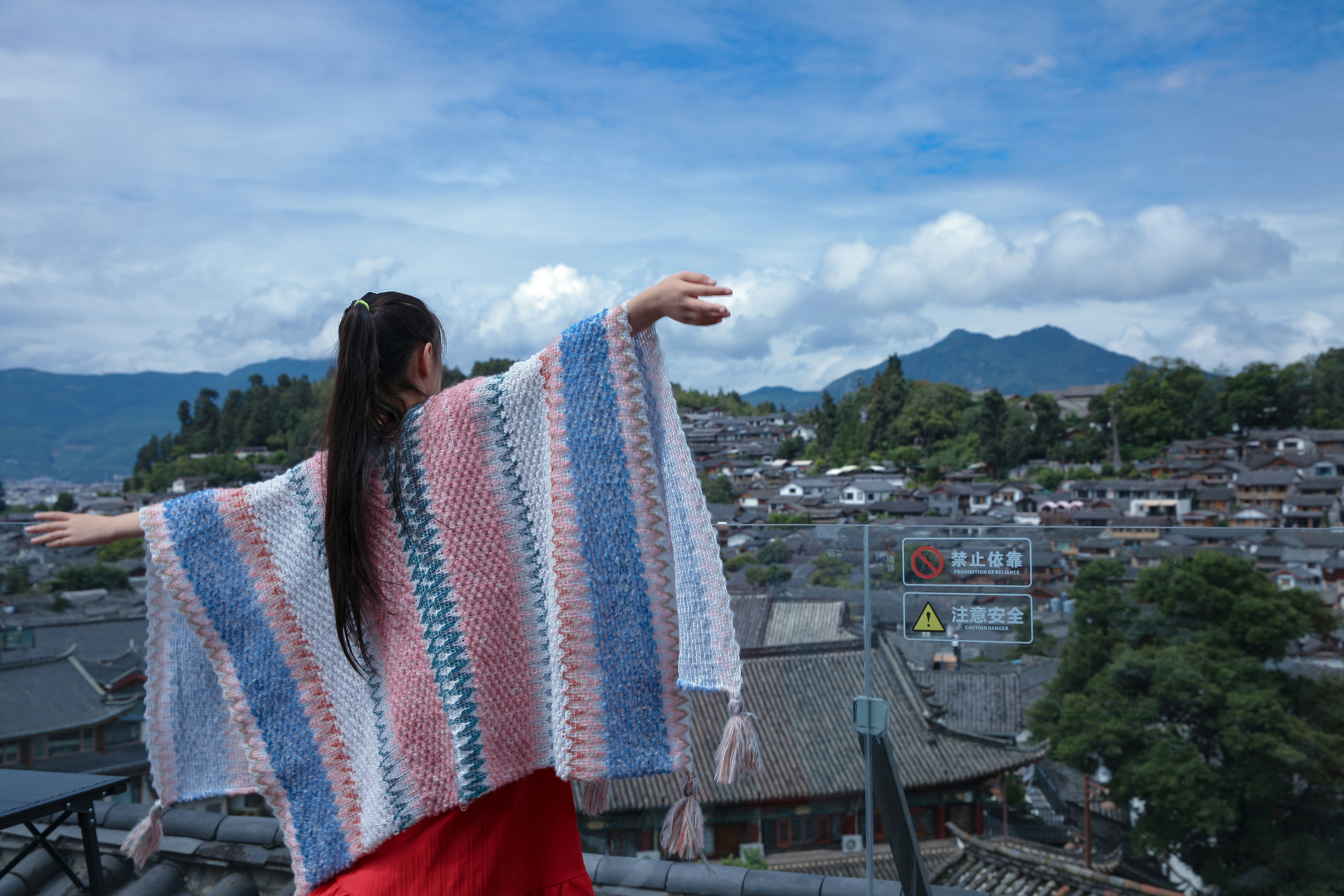Woman in a colorful shawl extends her arms towards the scenic mountainous landscape, overlooking a traditional village. Clouds drift across the sky.