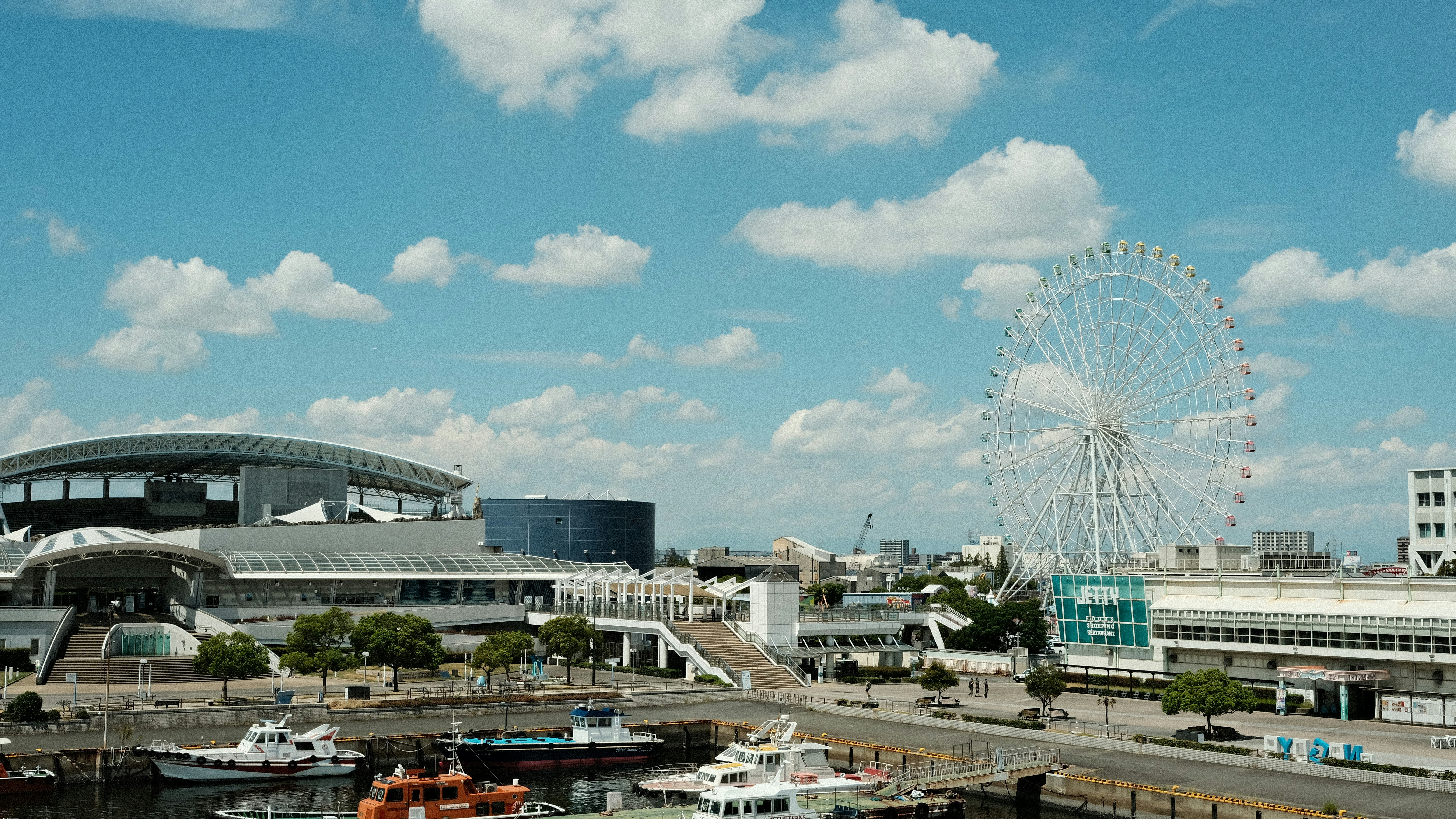 A vibrant waterfront scene featuring a Ferris wheel and modern architecture under a partly cloudy sky. Boats are docked nearby, creating a lively atmosphere.