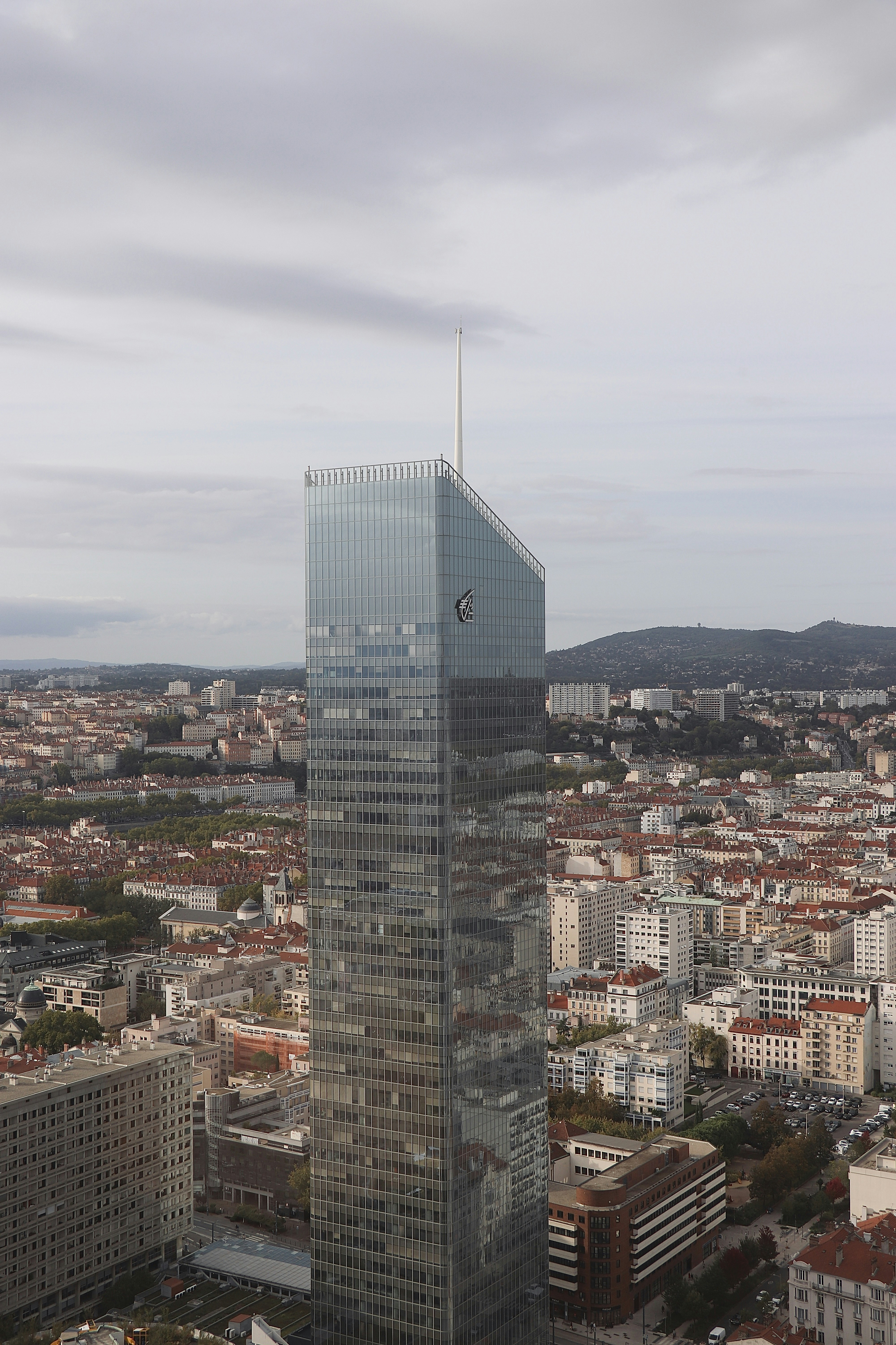 Aerial view of Lyon’s glass-clad Tour Incity rising above the Part-Dieu district, its spire cutting into a soft overcast sky. The reflective façade mirrors red-tiled rooftops and the city grid below, capturing the contrast between modern business towers and historic urban fabric. | Modern skyscraper dominates city skyline under cloudy sky