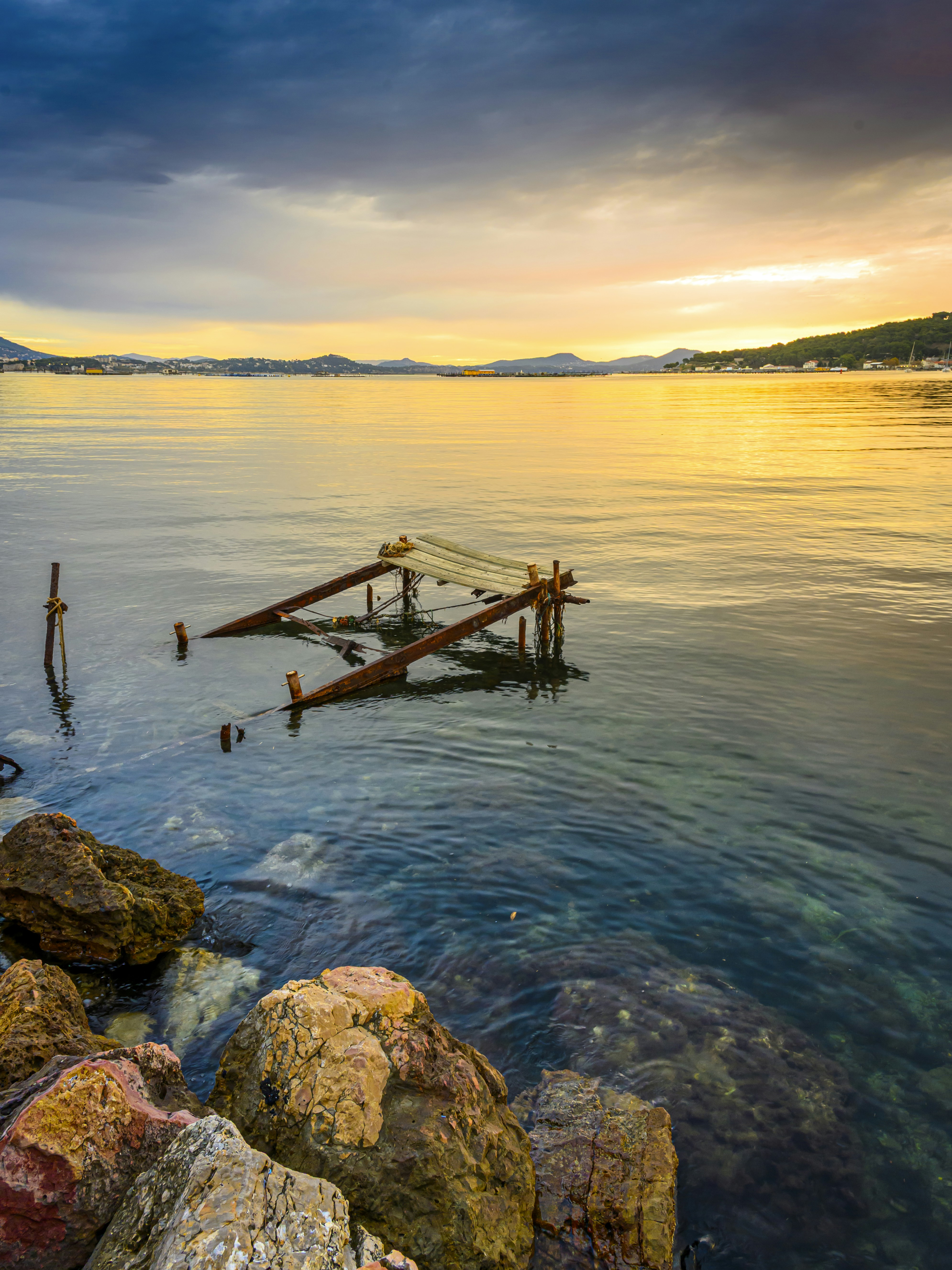 Abandoned wooden structure partially submerged in calm waters, framed by rocky shoreline and a golden sunset reflecting on the surface.