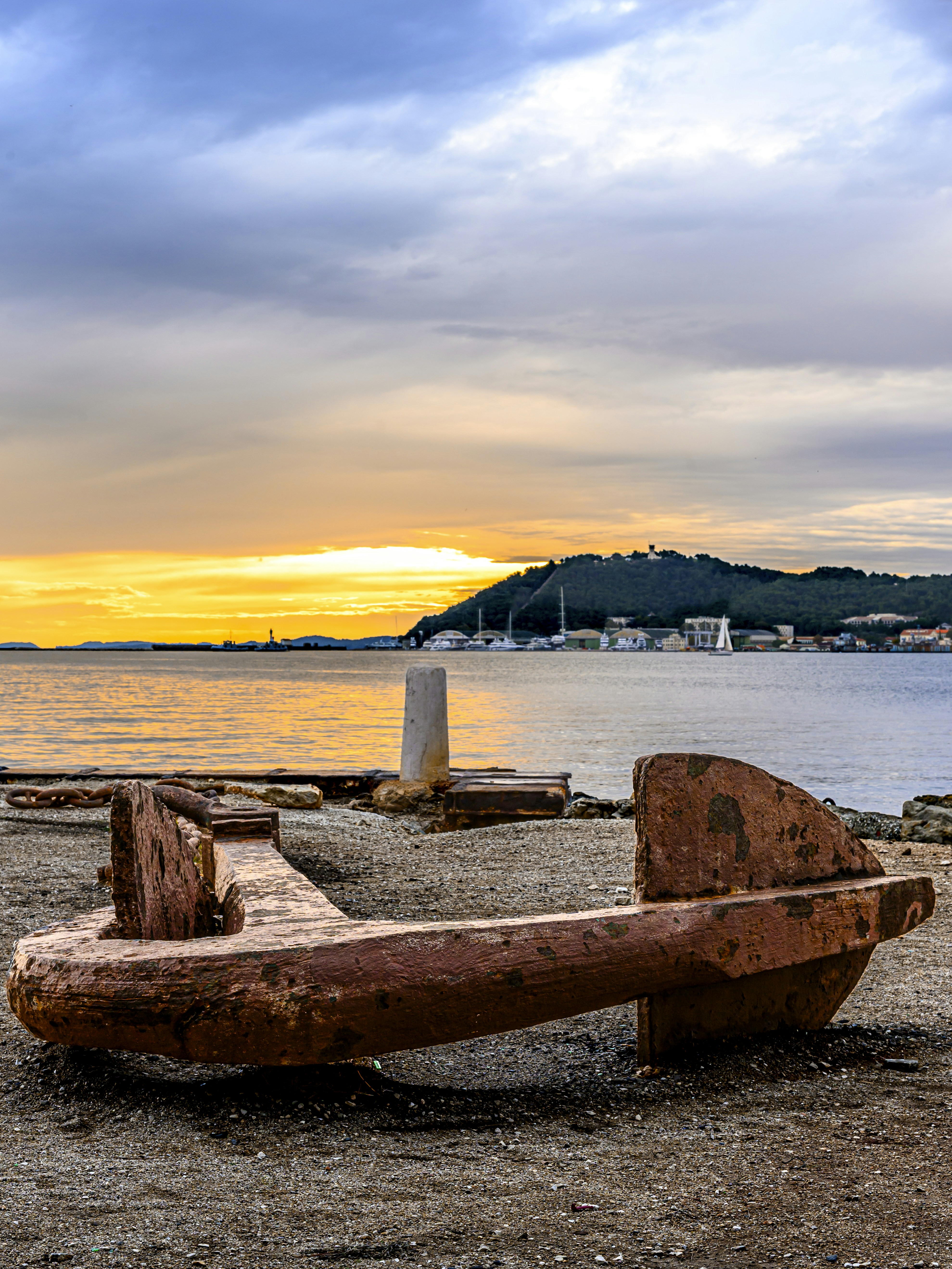 Weathered boat remnants resting on a pebbled shore, framed by a tranquil sunset over the water. The scene captures a moment of stillness and nostalgia.
