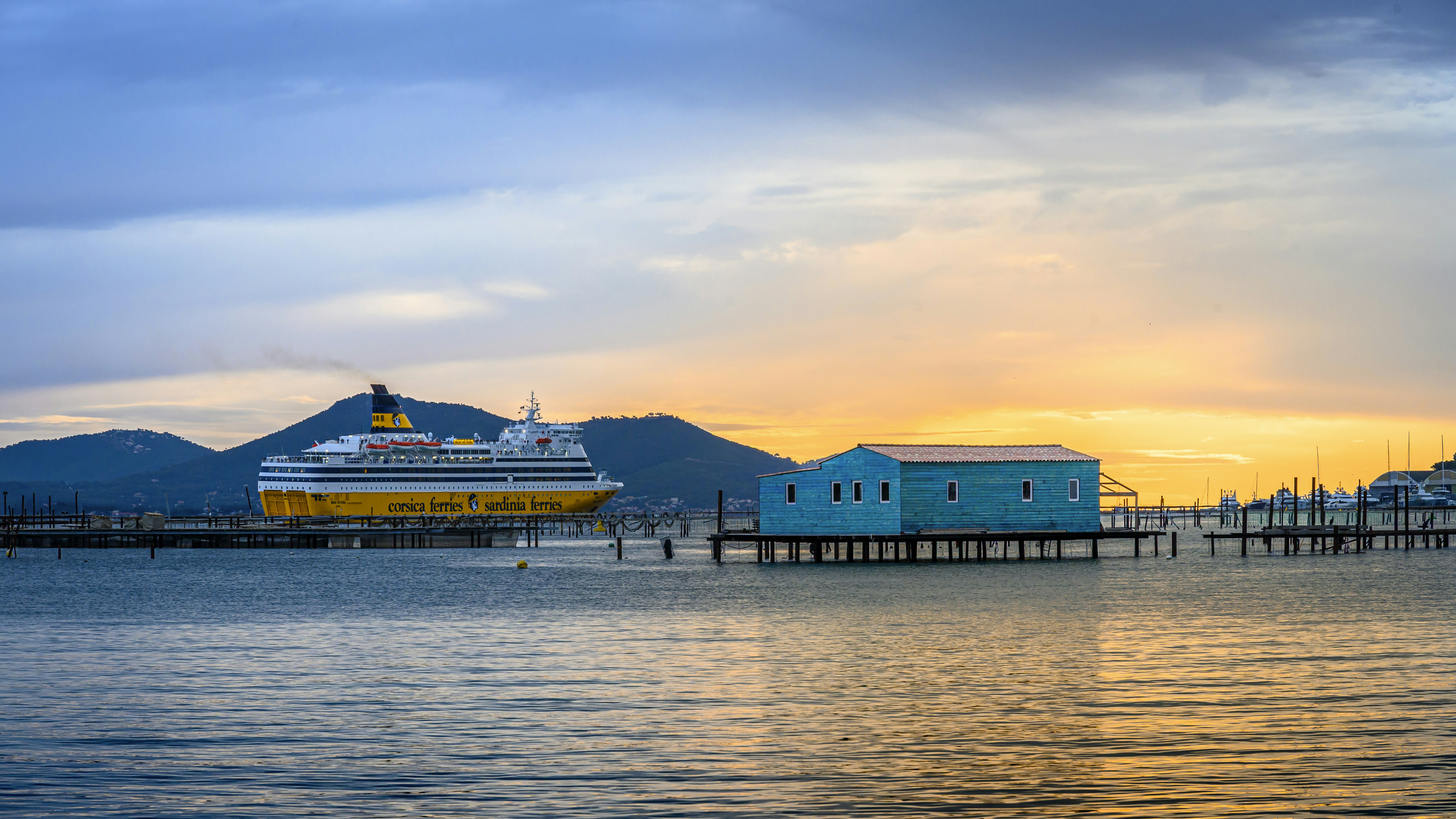 A ferry glides past a blue boathouse as the sun sets over a tranquil harbor, casting golden reflections on the water.
