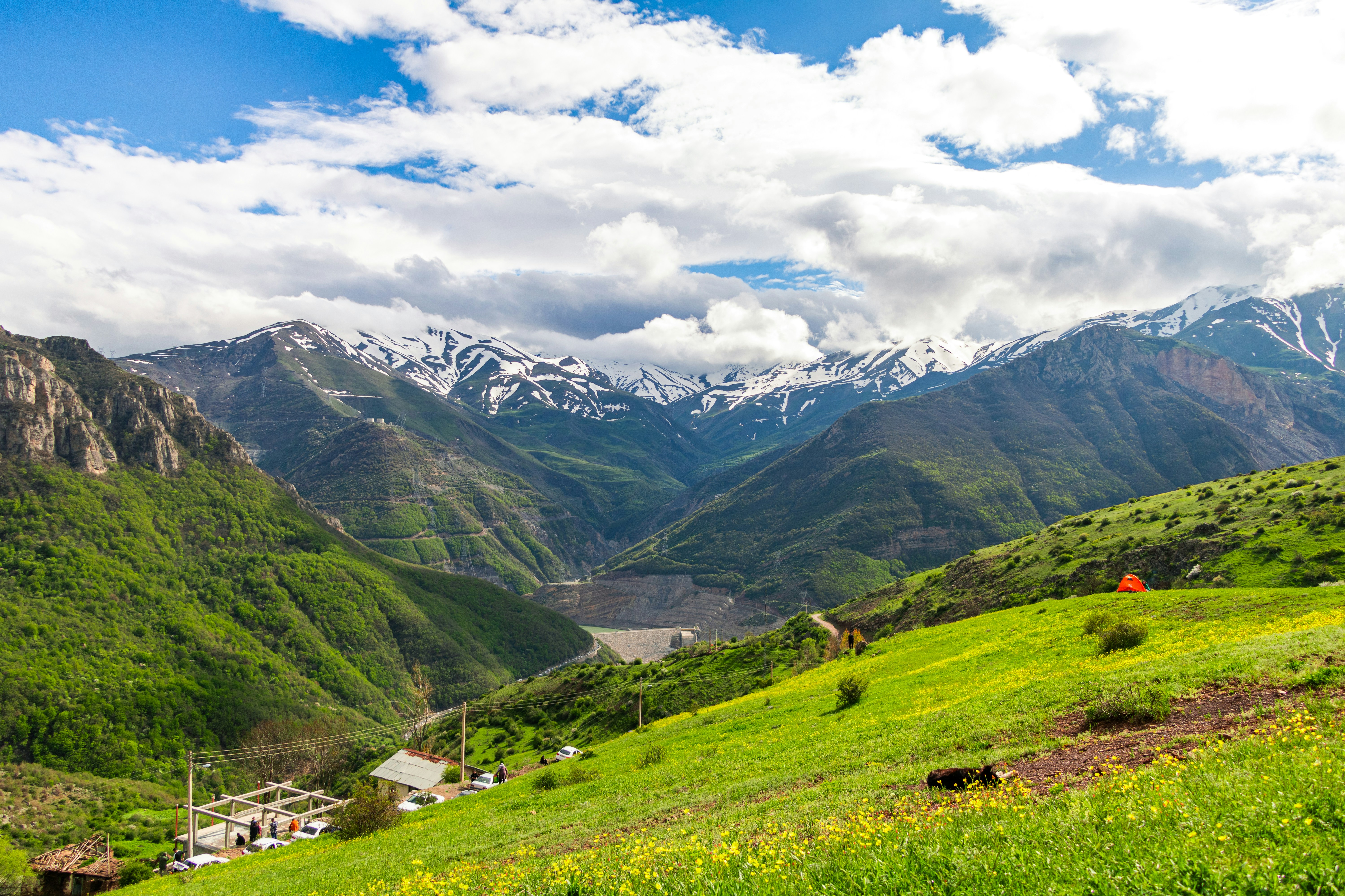 Lush green hillsides lead to a dramatic valley framed by towering, snow-capped mountains under a cloudy sky.