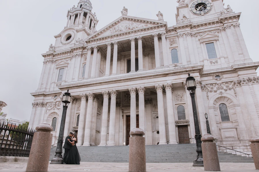 Couple in formal black attire embracing on the front steps of a grand neoclassical cathedral with tall columns and ornate stonework.