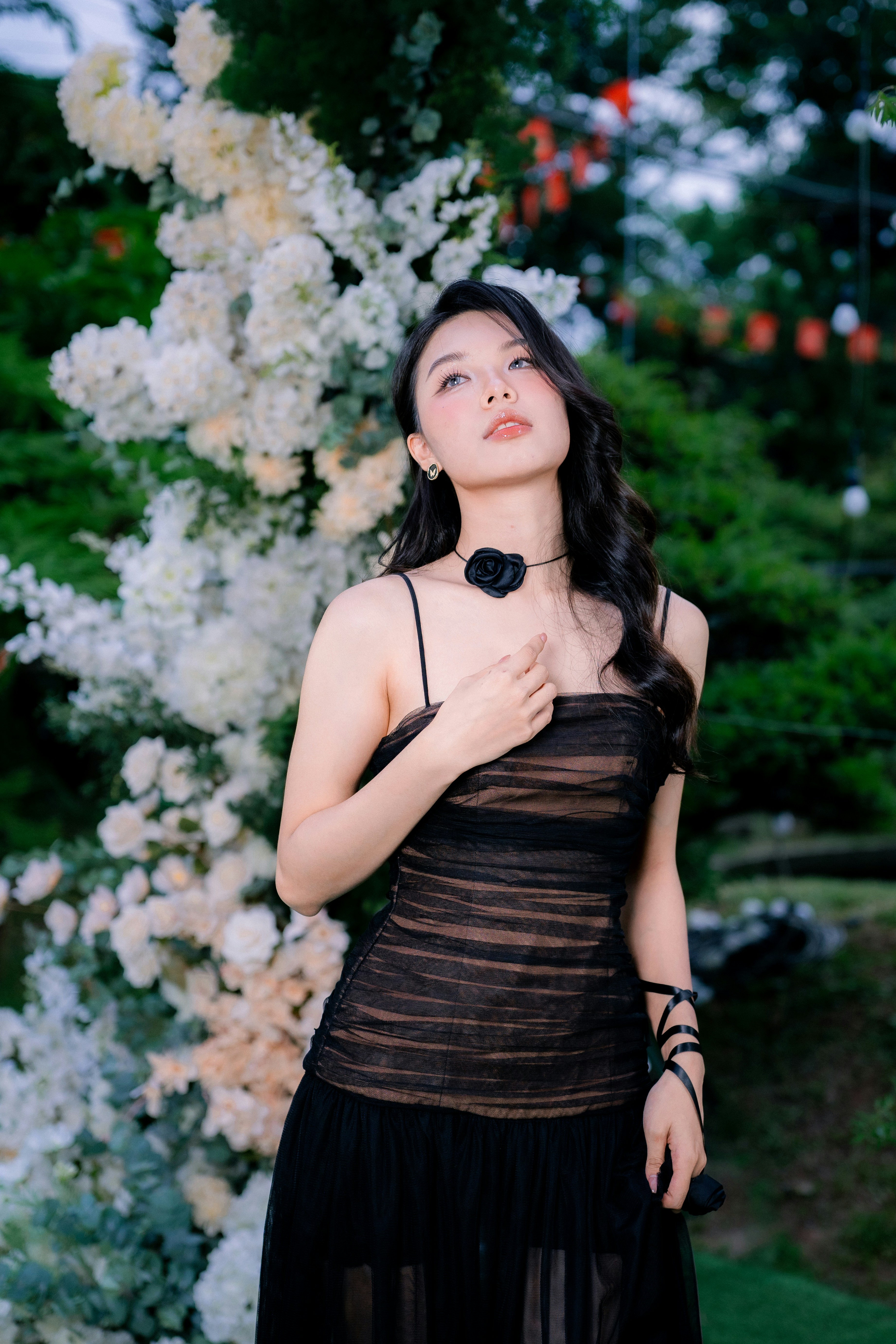 A young woman poses gracefully amidst a backdrop of lush greenery and delicate floral arrangements, exuding a sense of poise and tranquility.