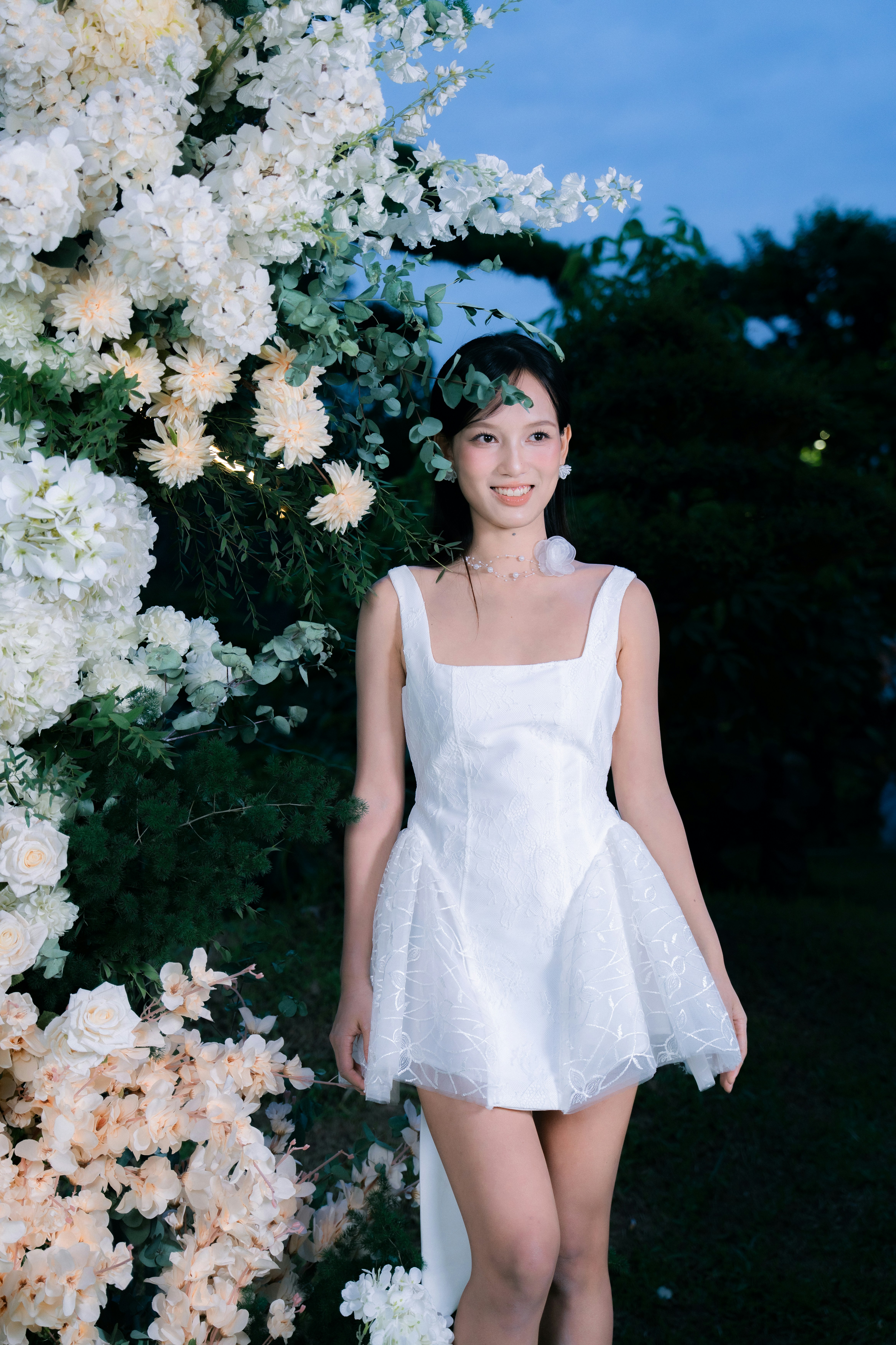woman in a white fashion dress | A woman in a white dress stands near floral decorations.