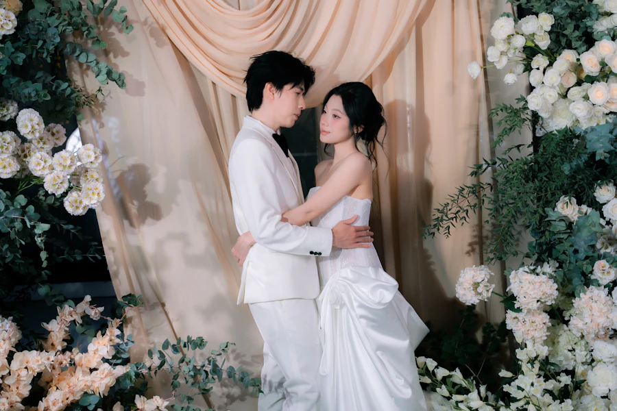 Bride and groom in white formalwear embracing before cream drapery and white floral arrangements. Soft, romantic stage decor frames the couple in a posed wedding portrait.