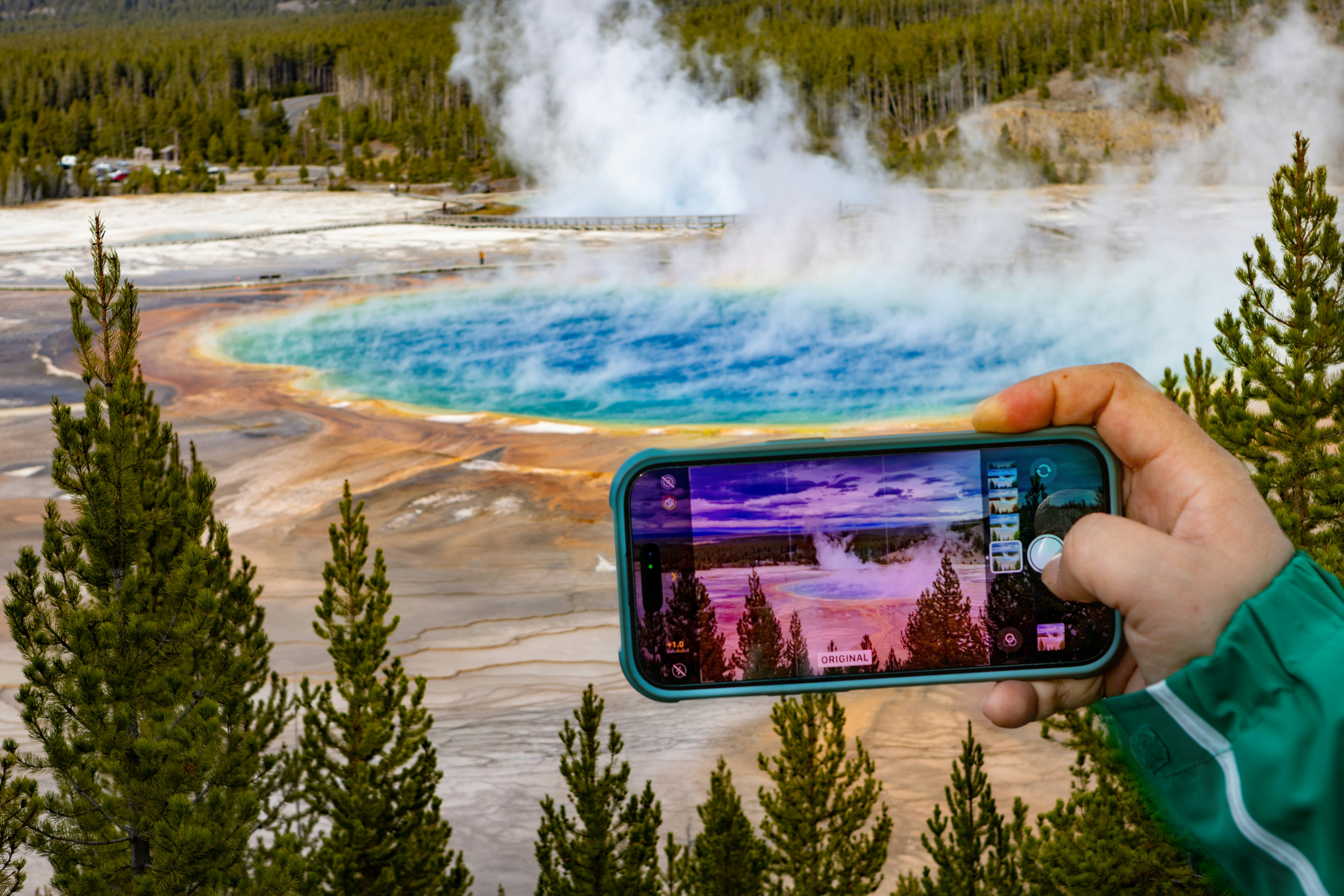 A hand holds a cellphone taking a photo of Grand Prismatic Spring in Yellowstone National Park on Saturday, October 25, 2025, as viewed from the obersvation viewing Platform on the Overlook Trail. | Hand holding phone photographing colorful hot spring