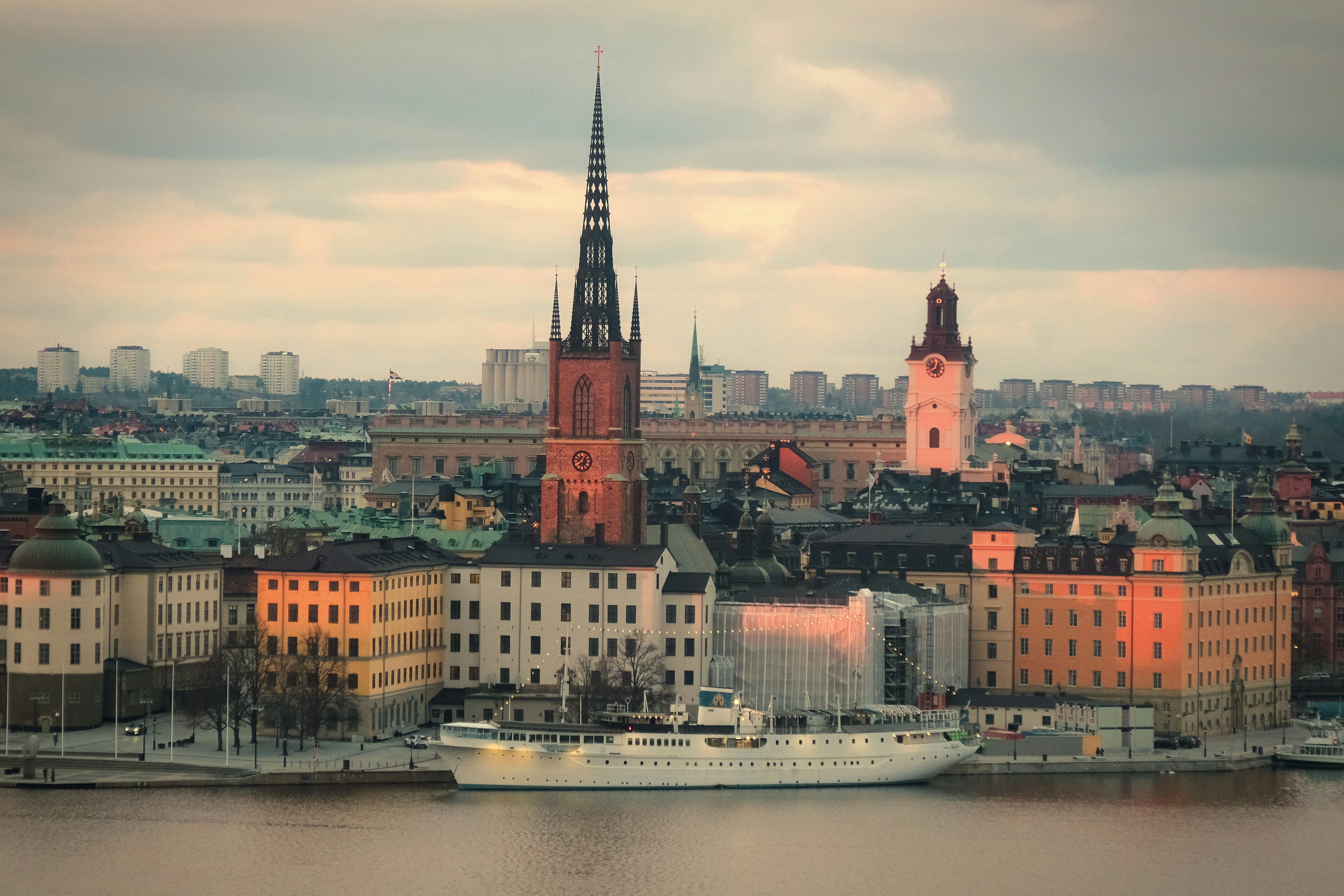 Scenic cityscape of Stockholm, Sweden, captured from Skinnarviksberget, the city’s highest natural viewpoint. The image highlights Riddarholmen Church with its iconic spire, historic architecture, and waterfront buildings at sunset. | Historic cityscape with a tall spire and ship.