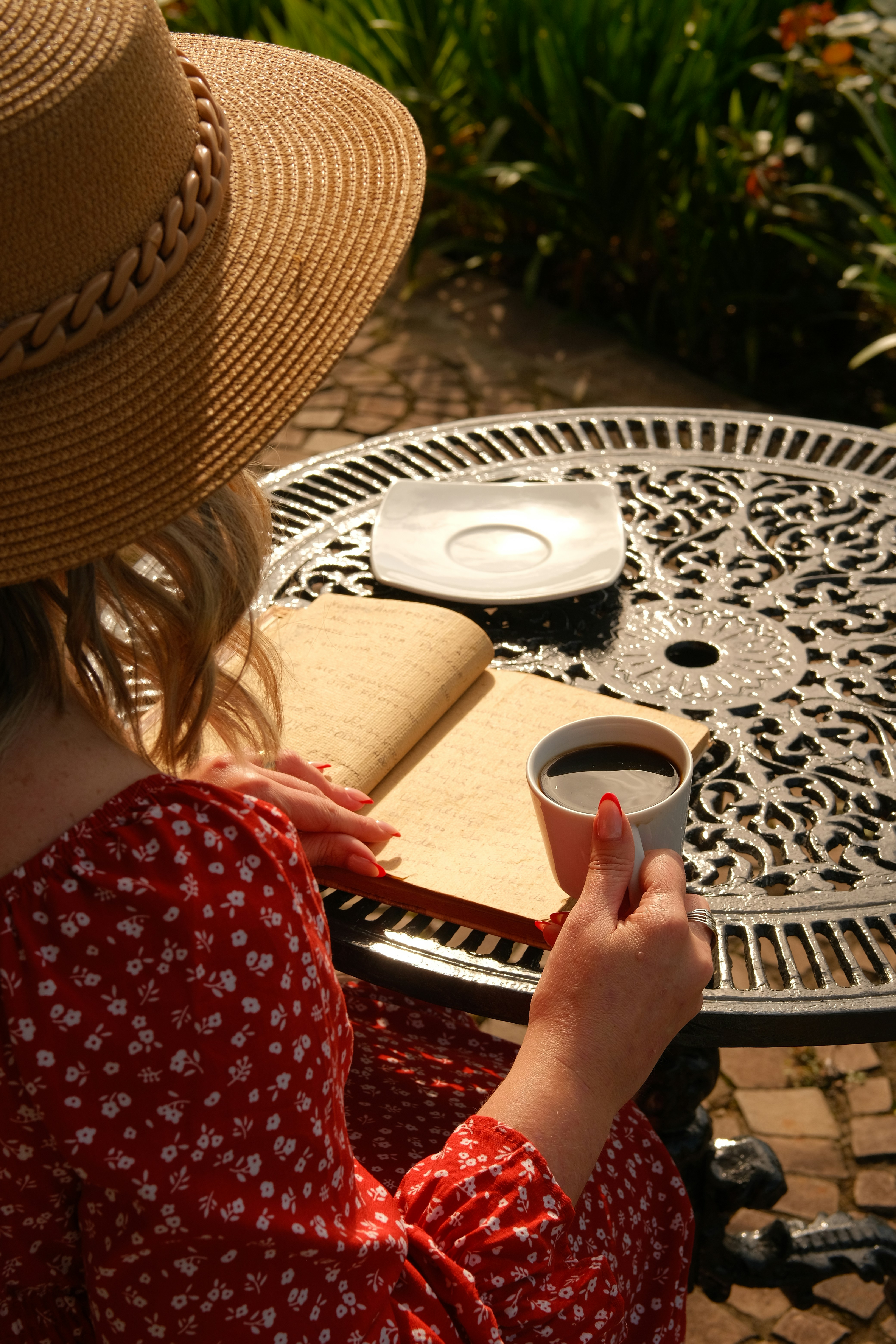 Woman in a floral dress enjoying coffee while reading a book at a garden table. Sunlight casts warm tones on the scene.