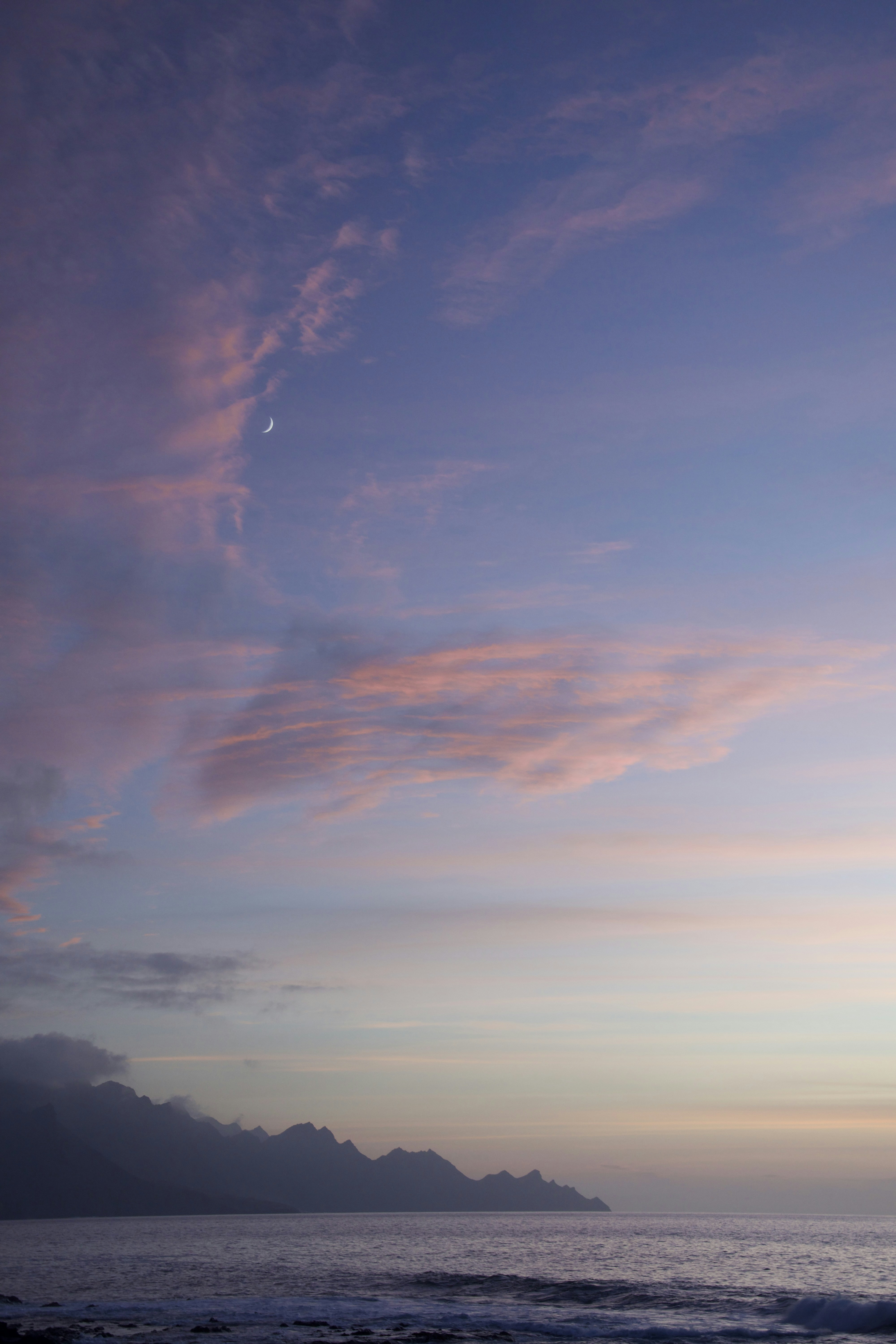 Soft pastel clouds stretch across the sky as a crescent moon rises above a tranquil ocean view, framed by distant mountains.
