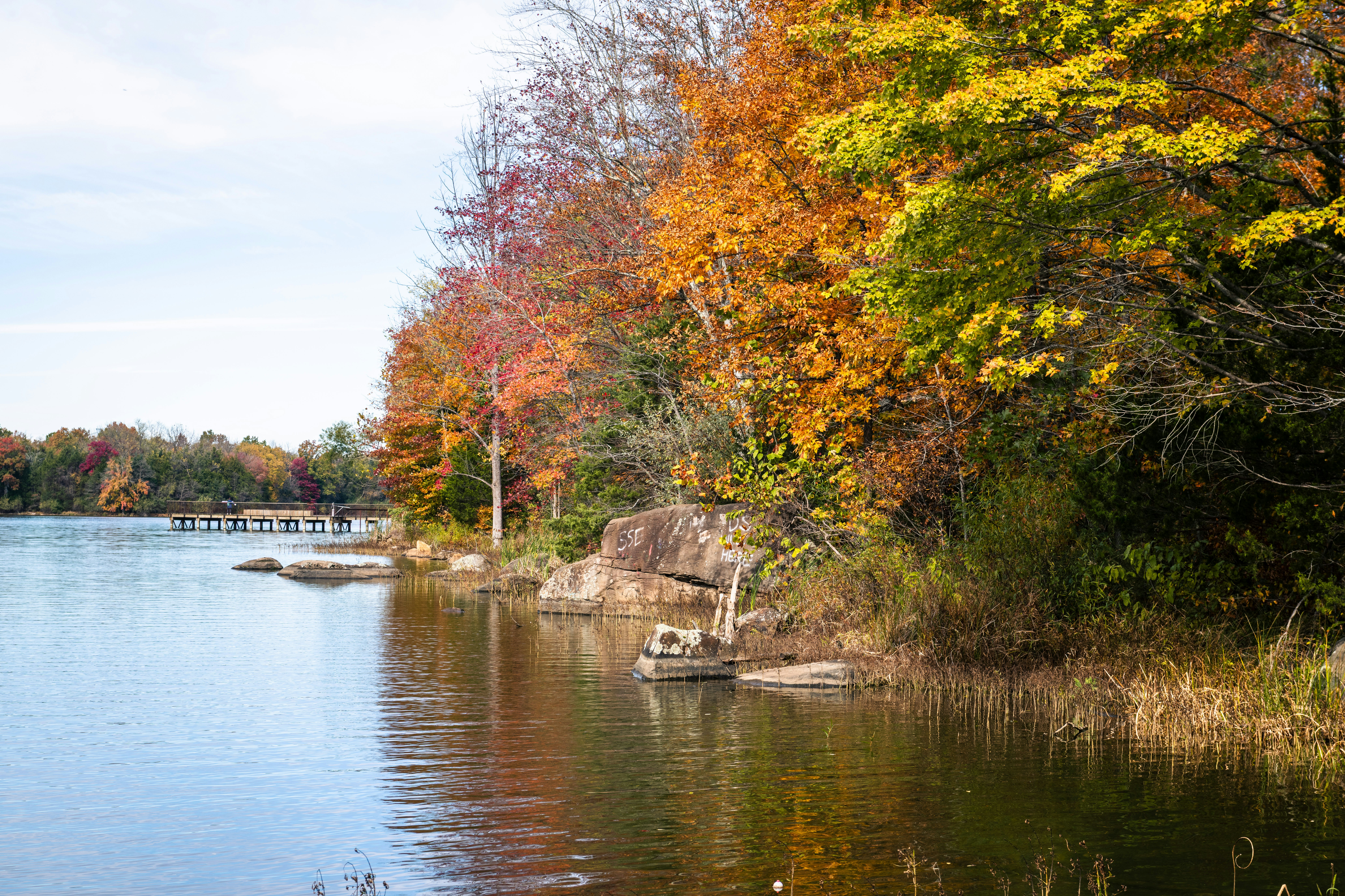 Vibrant autumn foliage reflects on calm waters beside a rocky shoreline, creating a serene landscape. A wooden pier is visible in the distance.