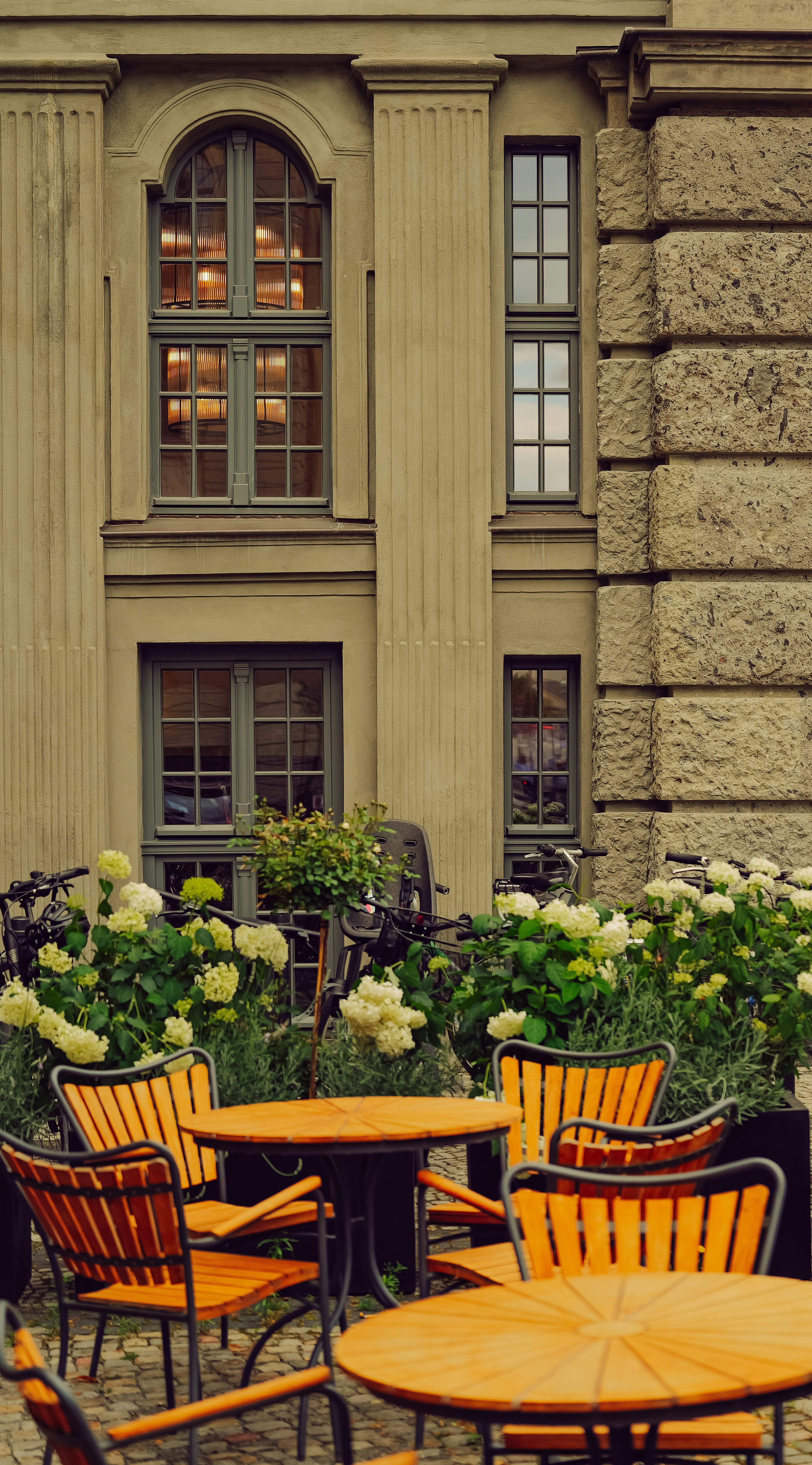 Charming outdoor café scene featuring vibrant orange tables surrounded by lush greenery and bicycles, set against a historic building facade.