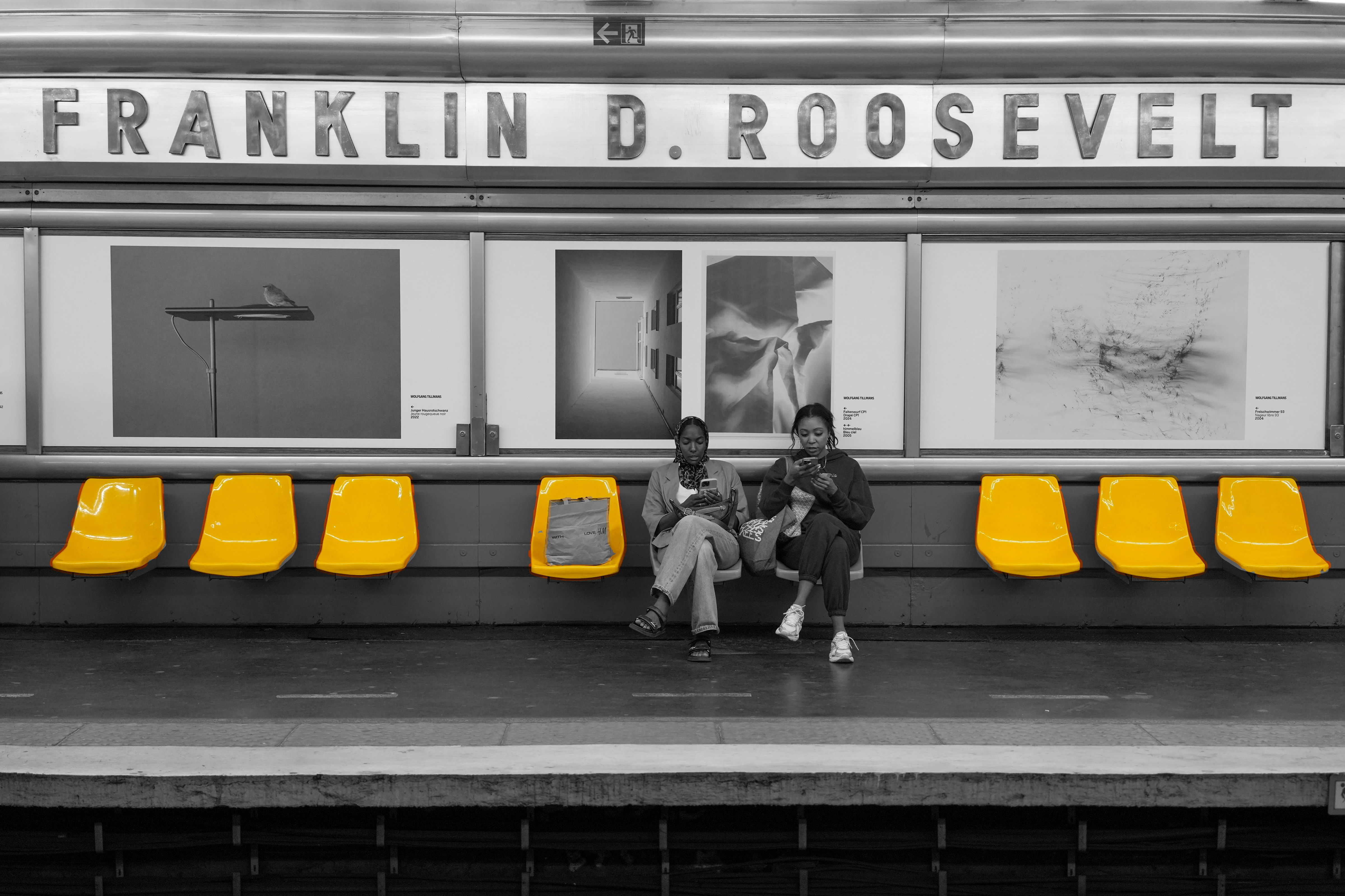 Two people sit on yellow subway seats under station sign.