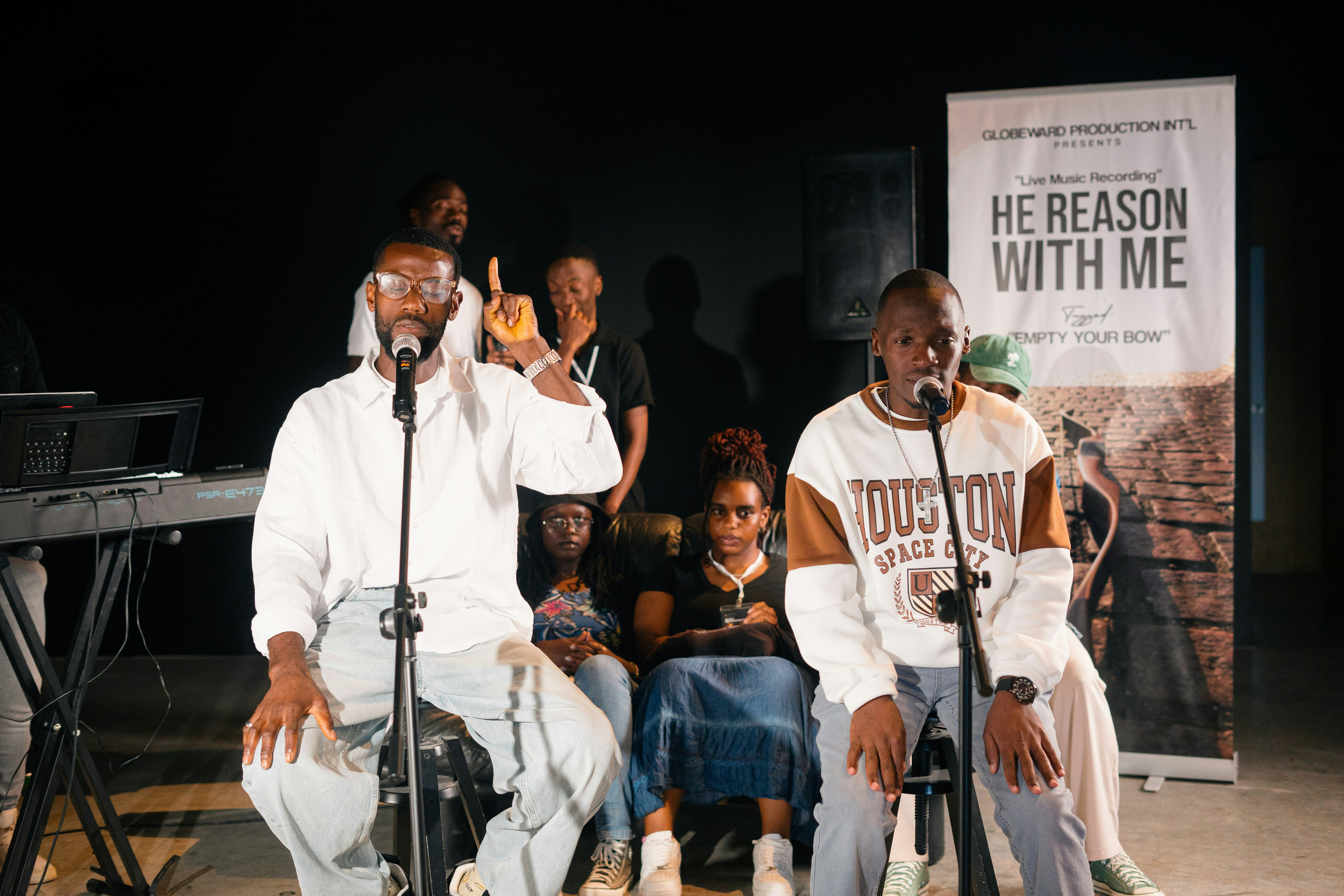Two speakers engage in a heartfelt discussion, while an attentive audience listens in a dimly lit setting. A promotional banner for the event is visible in the background.