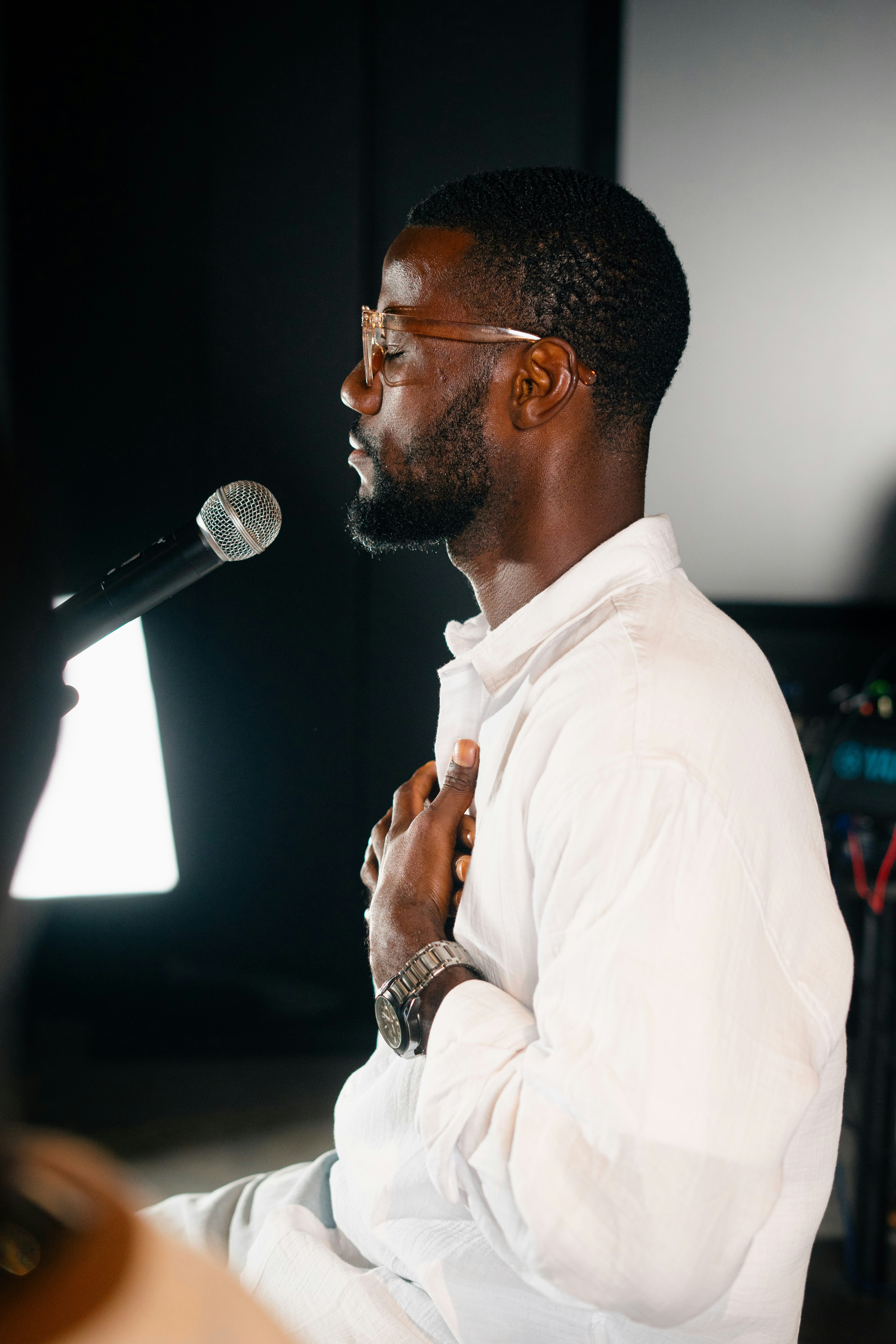A man in a white shirt deeply engaged in thought while seated near a microphone, illuminated by soft lighting.
