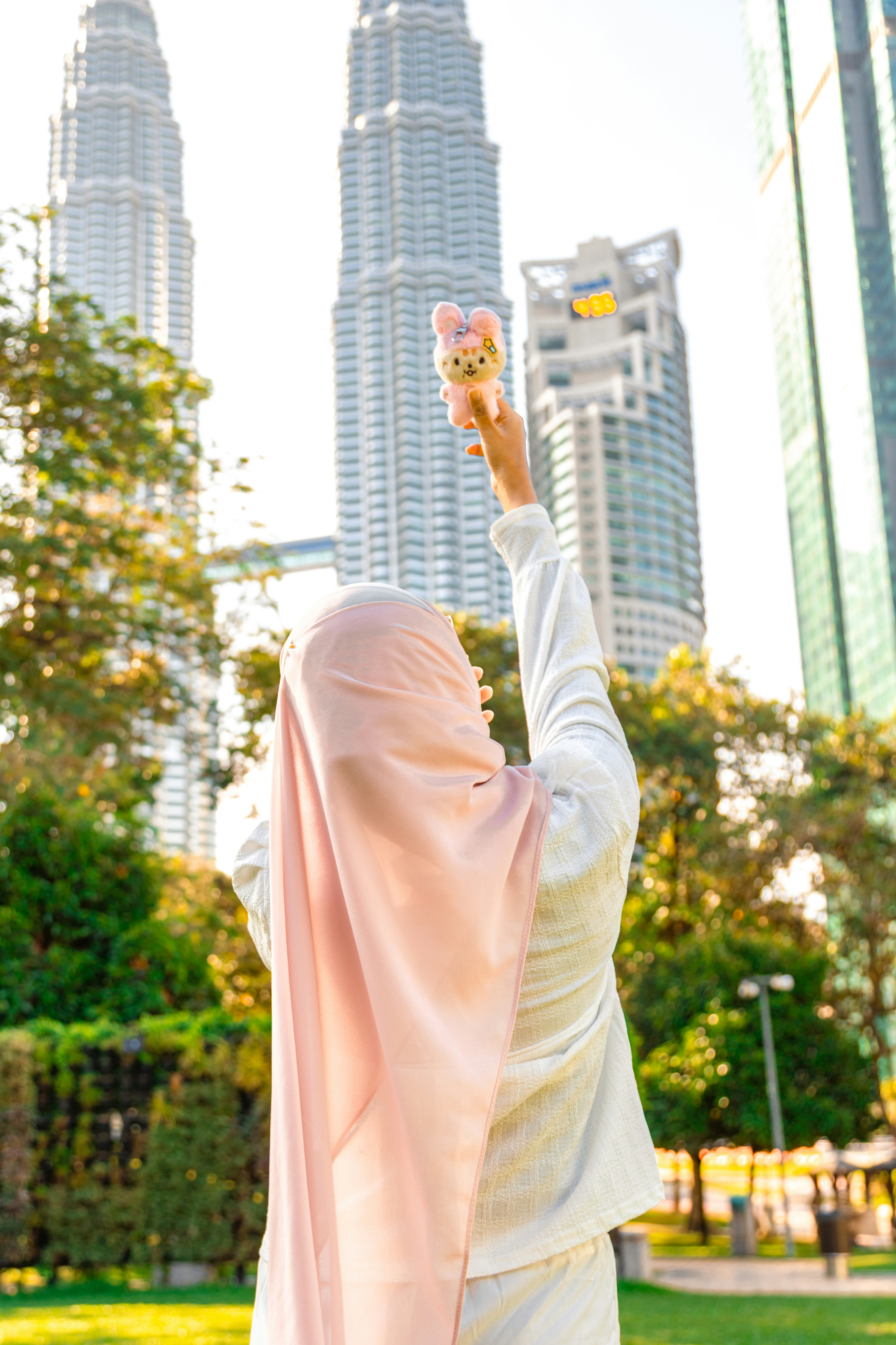 A woman in a pink hijab holds a plush toy high against the backdrop of towering skyscrapers, celebrating a moment of joy in a vibrant urban park.