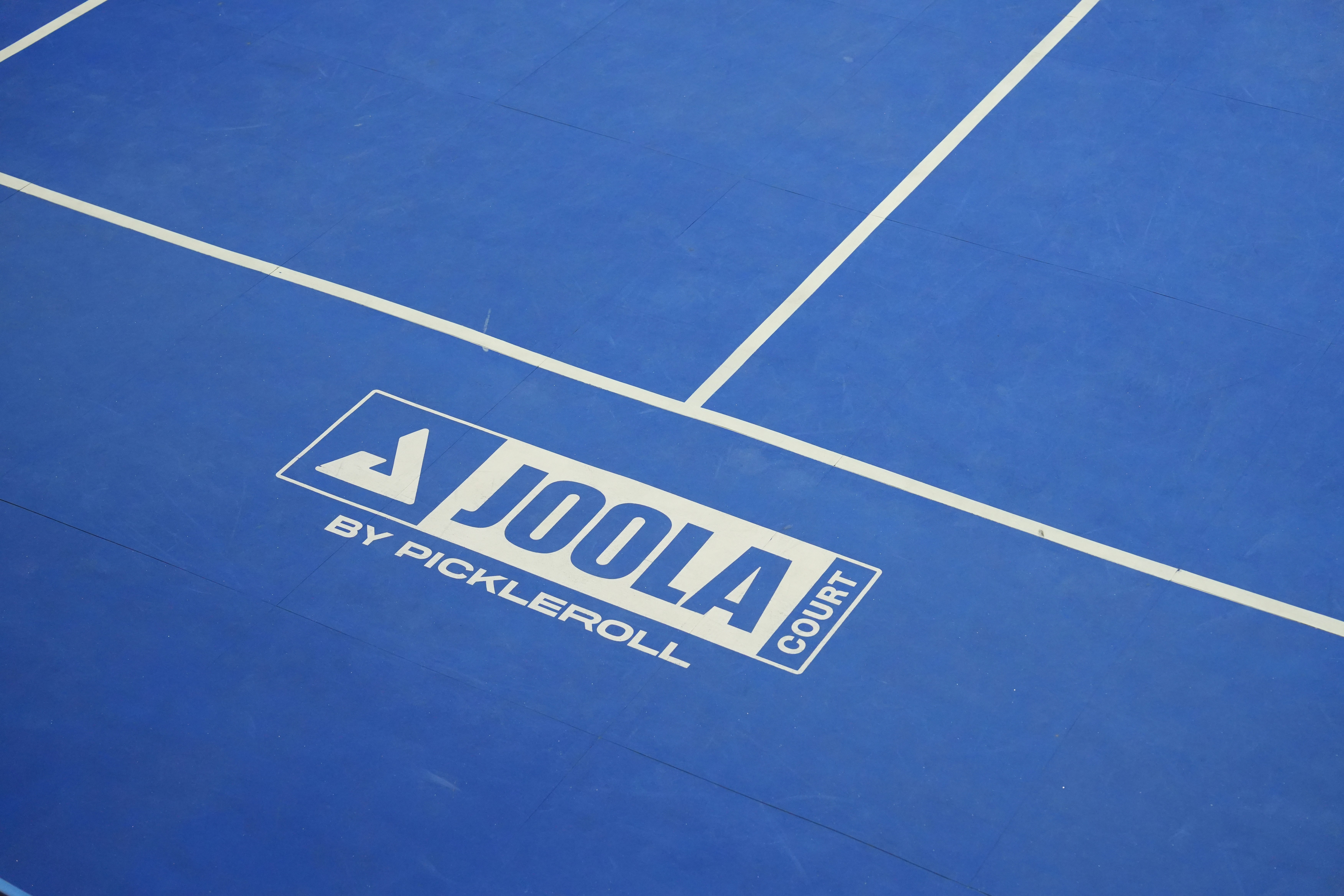 Bright blue pickleball court featuring the Joola logo prominently displayed. The court's vibrant color enhances the dynamic atmosphere of the sport.
