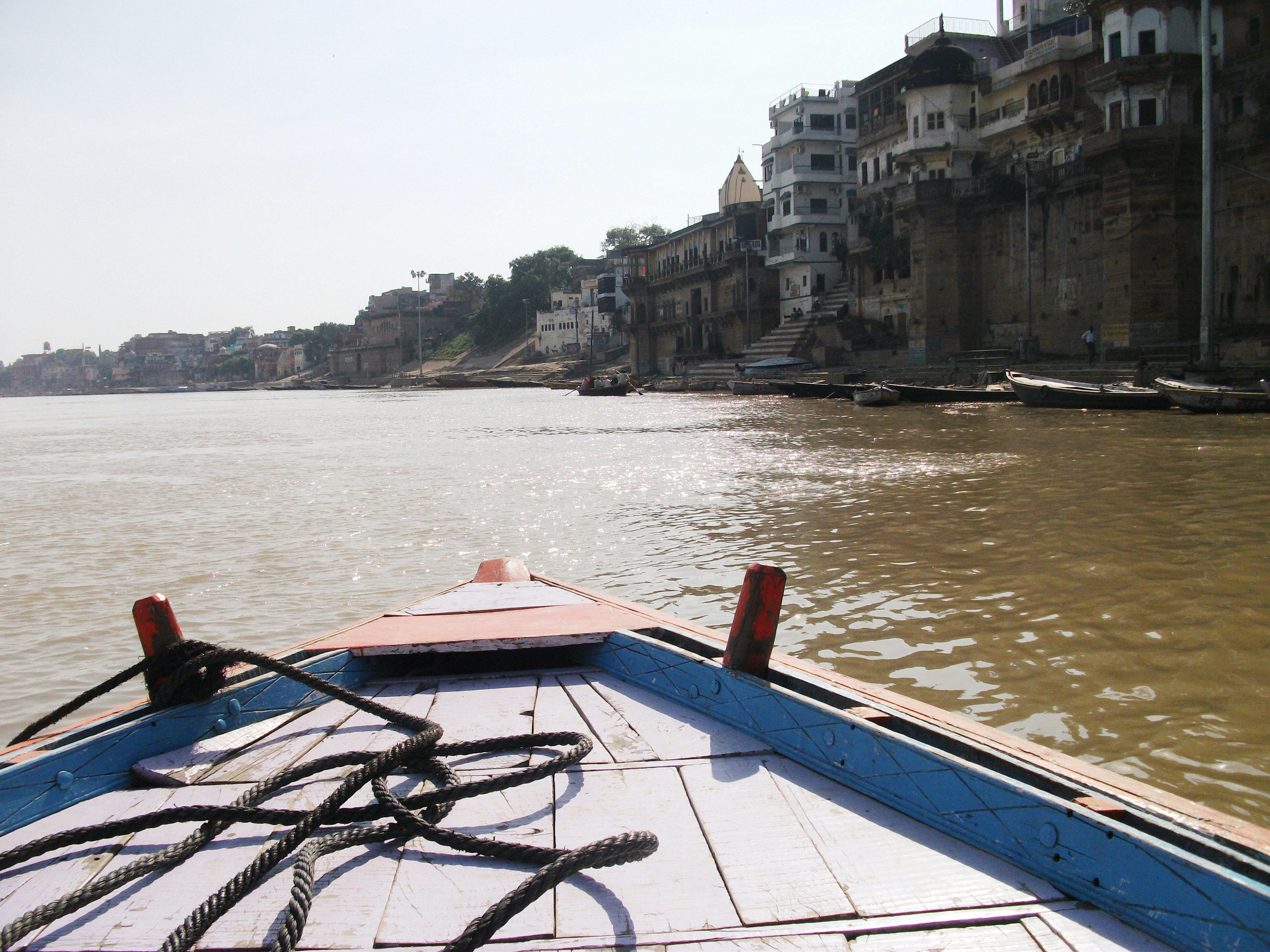 Ganga Devi, The Mighty Godess Force of Sacred Water from Heaven to Earth. To Control Her Flow Smoothly to Earth The God of Destroyer Shiva, kept Ganga Devi on his Head and Opened a Small Hair Lock in Which the Flow of Ganga Devi was controlled smoothly to earth. Ganga Devi is considered the Holy River of Earth in its Purest Form of Water with its Magnetic Mysterious Energy. Ganga Devi Harati is given to River Goddess Ganga Every Day Evening on the Ghats of Varanasi. | Boat moving along a river with buildings on shore.