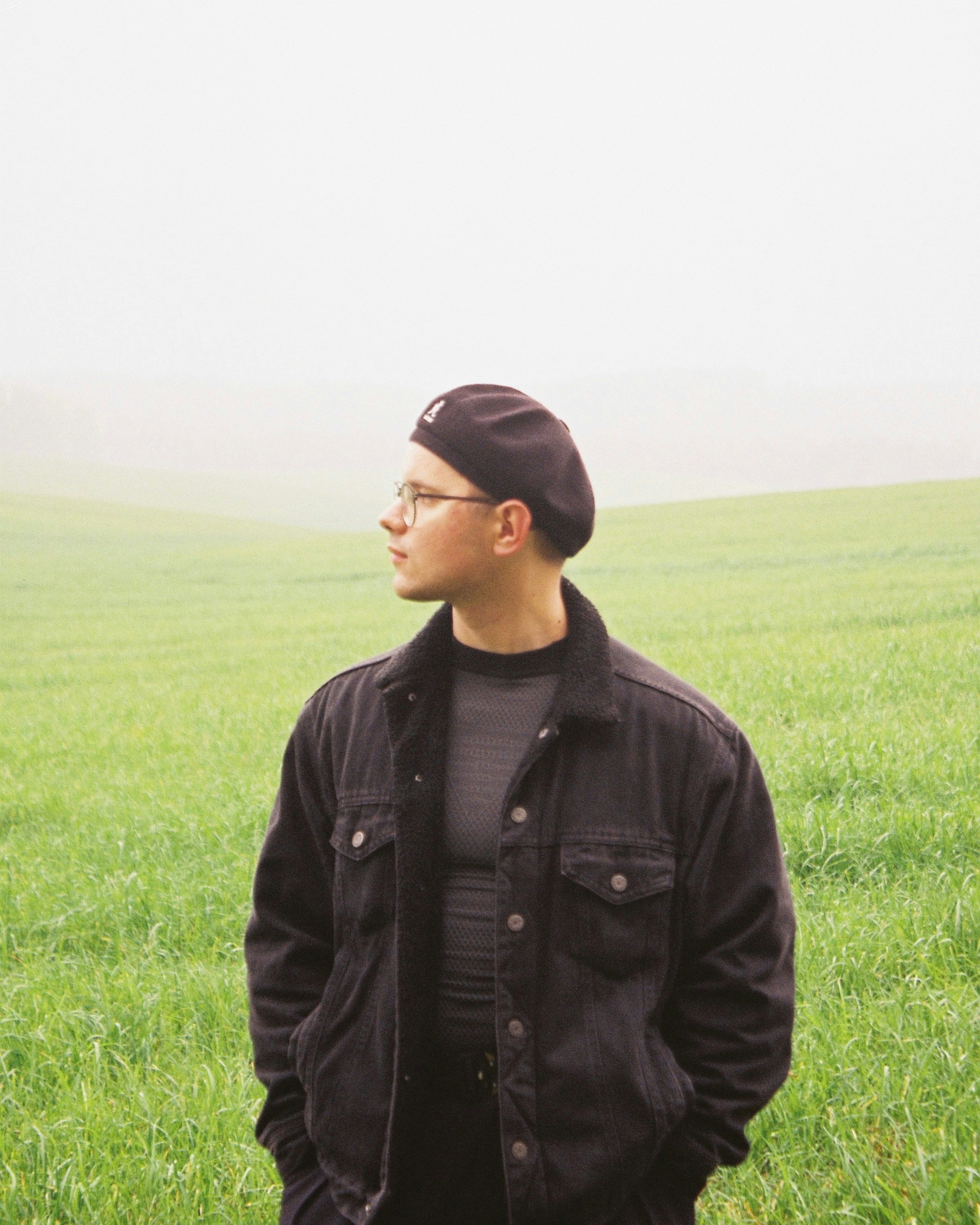 Individual in a black jacket and beret stands contemplatively in a foggy green field, surrounded by soft, muted landscapes.