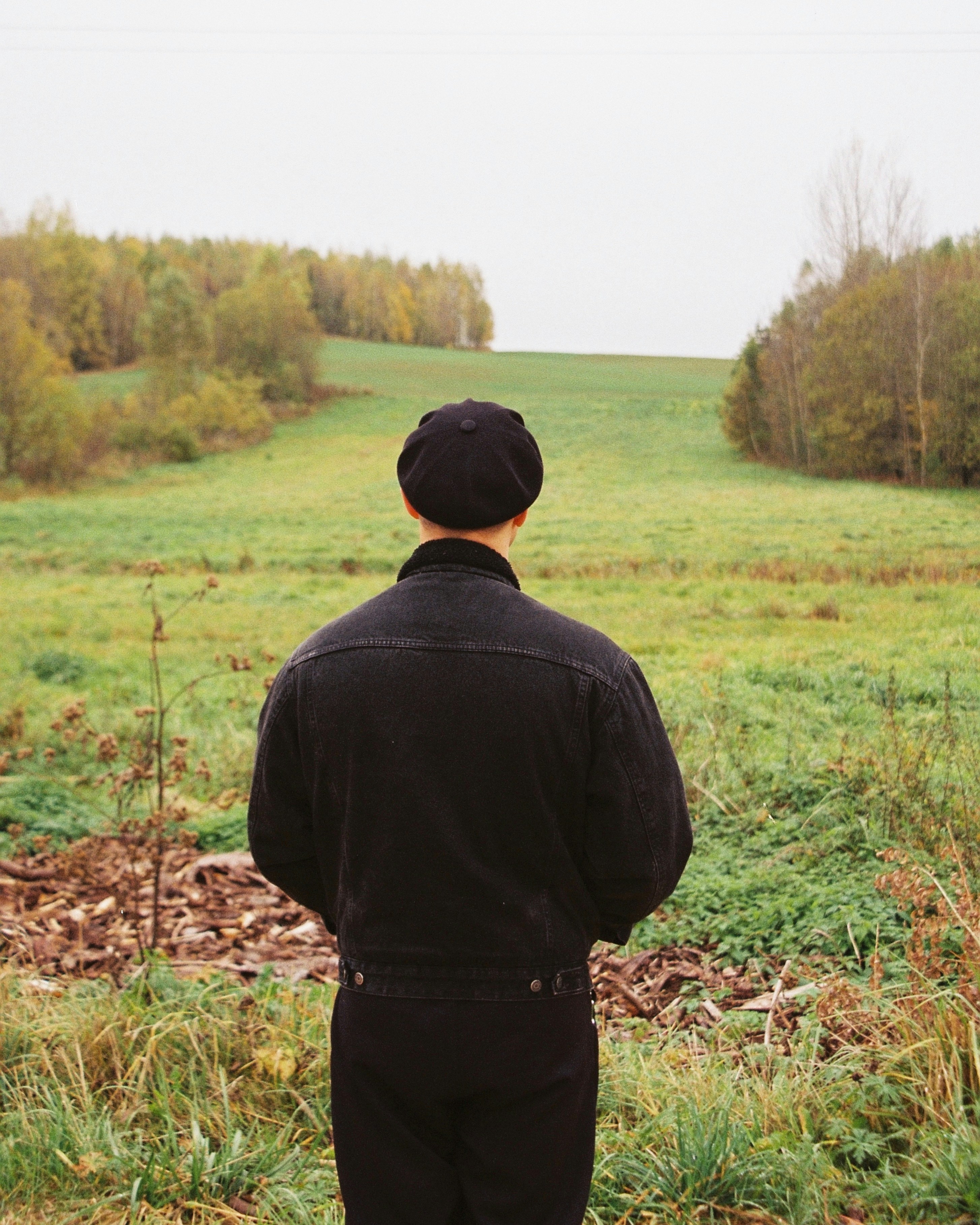 Individual in a black jacket and beret gazes over a lush green field, surrounded by trees in autumn colors.