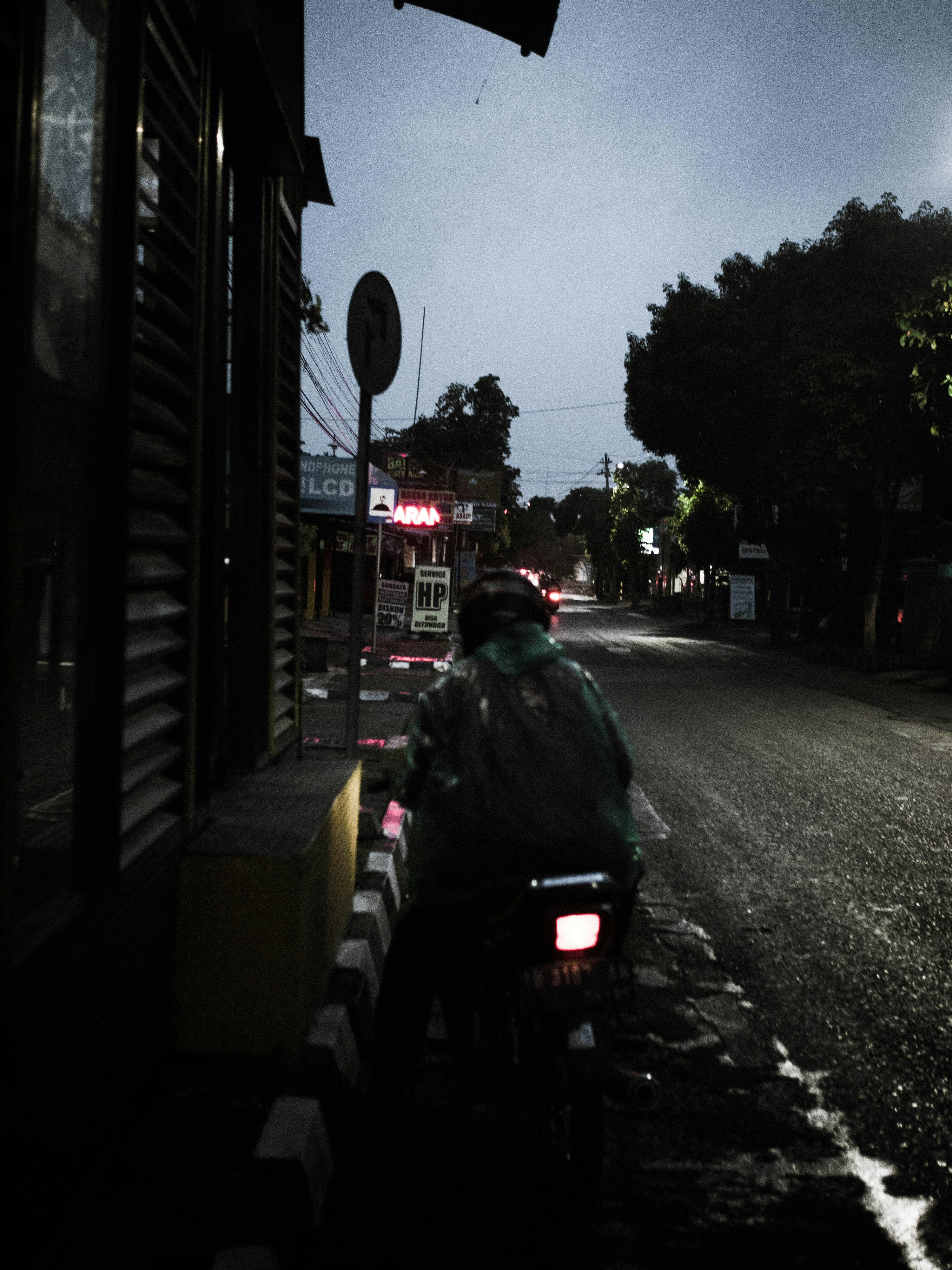 A motorcyclist clad in a raincoat navigates a dimly lit street, surrounded by reflections and shadows of urban life.
