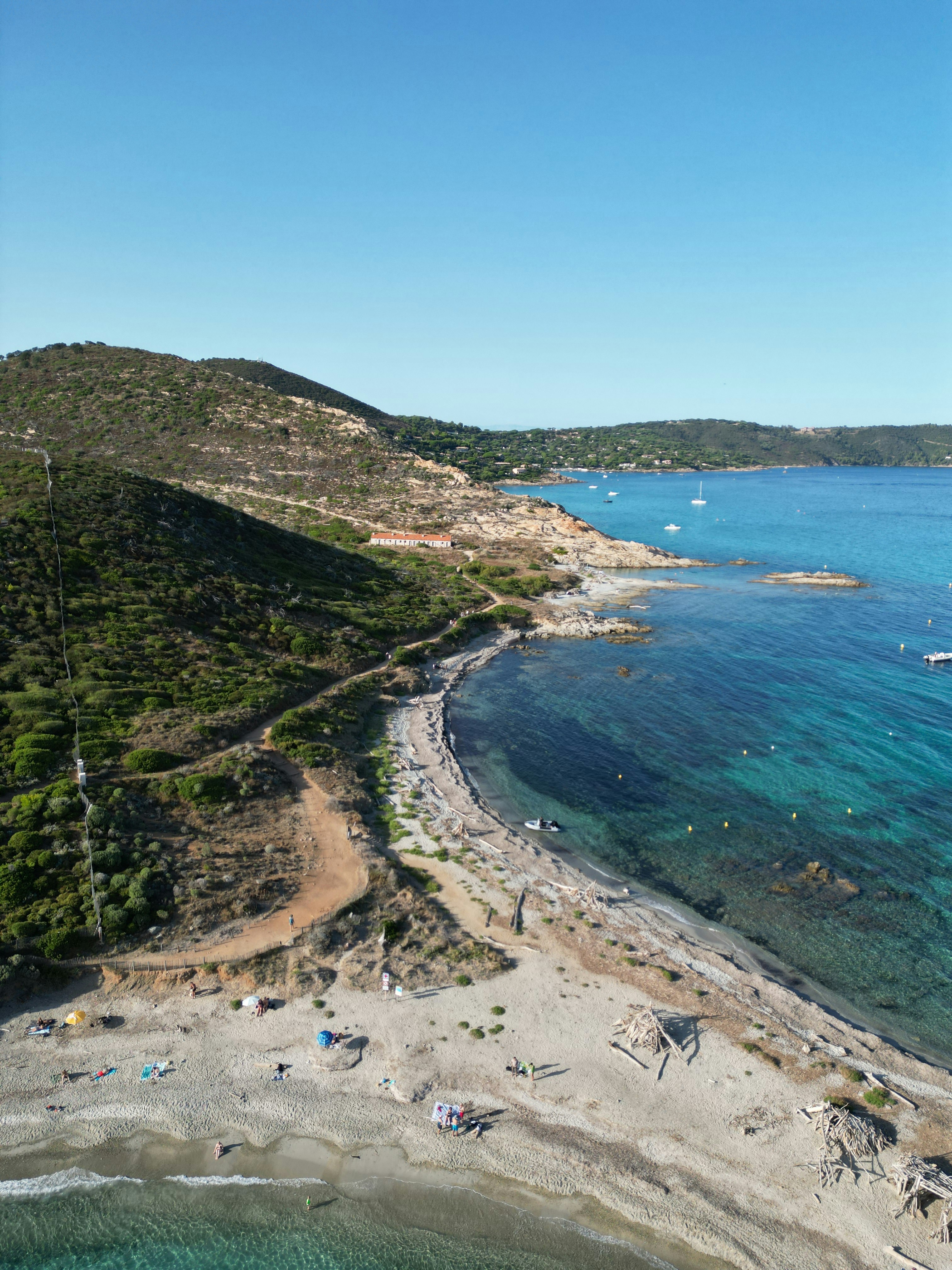 Aerial view of a tranquil beach bordered by lush hills and clear blue waters, showcasing boats and beachgoers enjoying the sun.