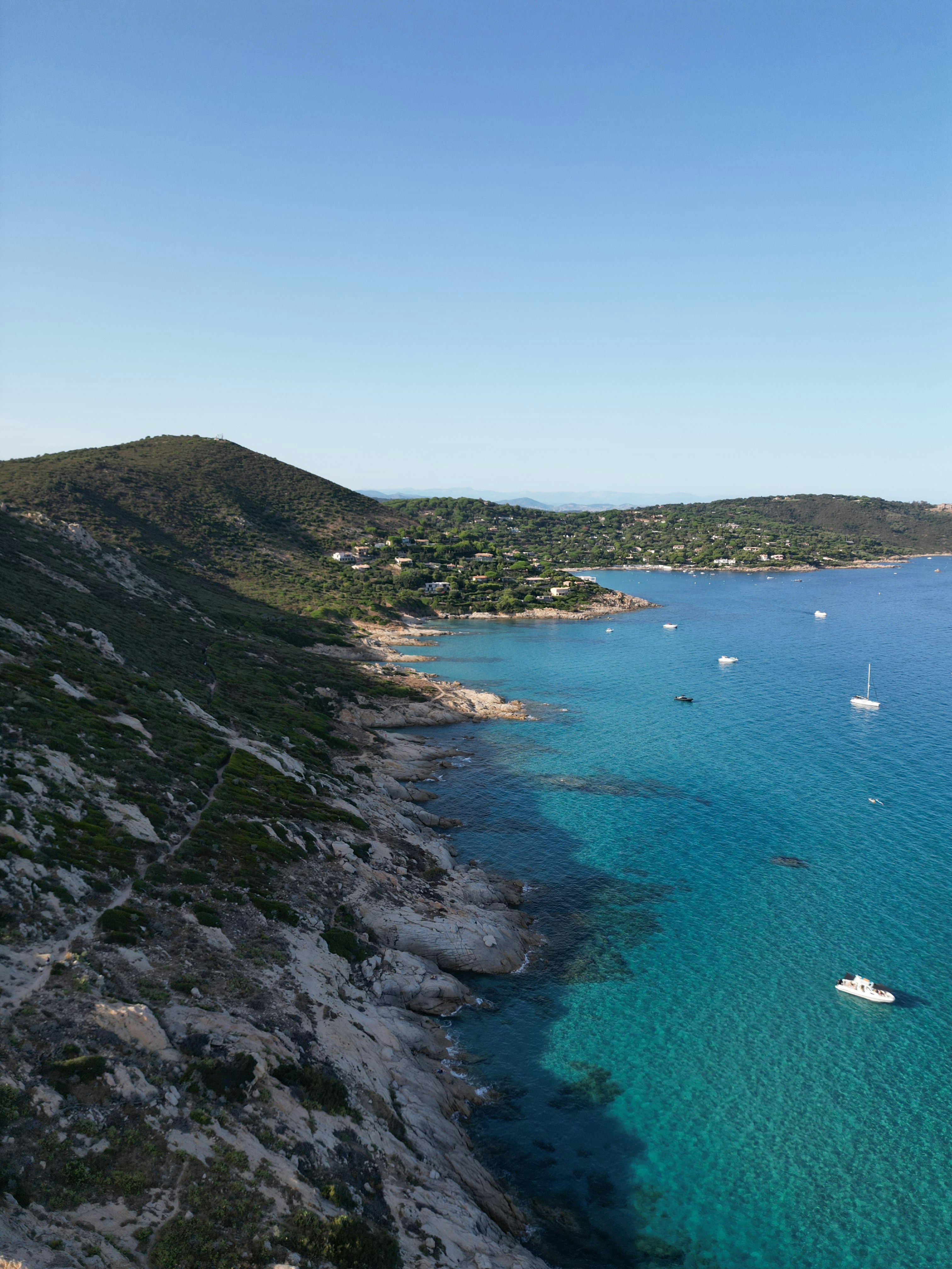 Sailboats anchored in a clear blue bay near rocky coast.