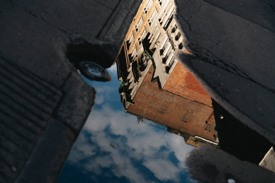 A brick apartment building and cloudy blue sky reflected in a puddle framed by dark pavement. The distorted reflection creates an abstract urban scene.