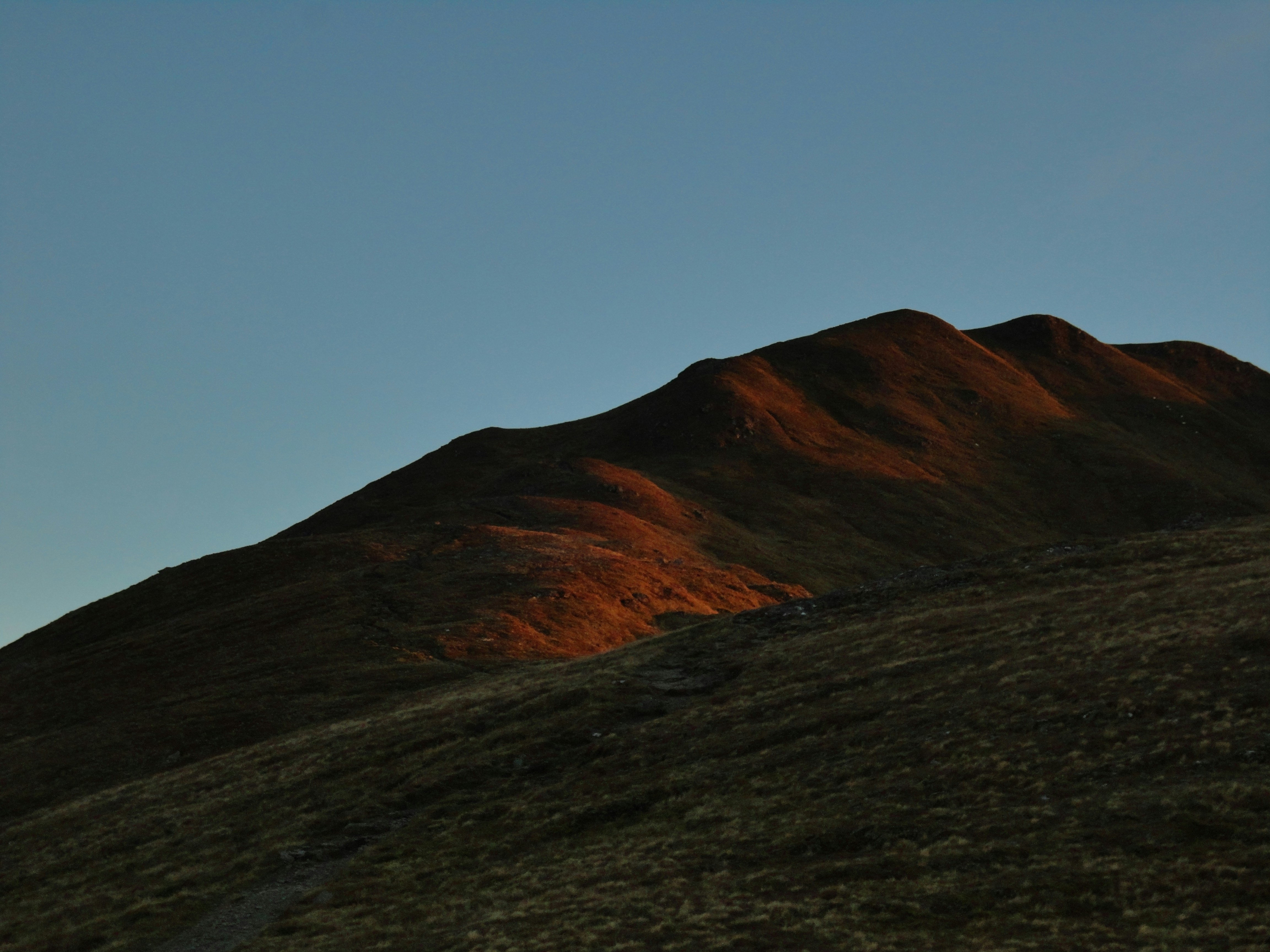 Sunset light gently caressing the contours of a mountain, revealing its rugged texture against a clear sky.