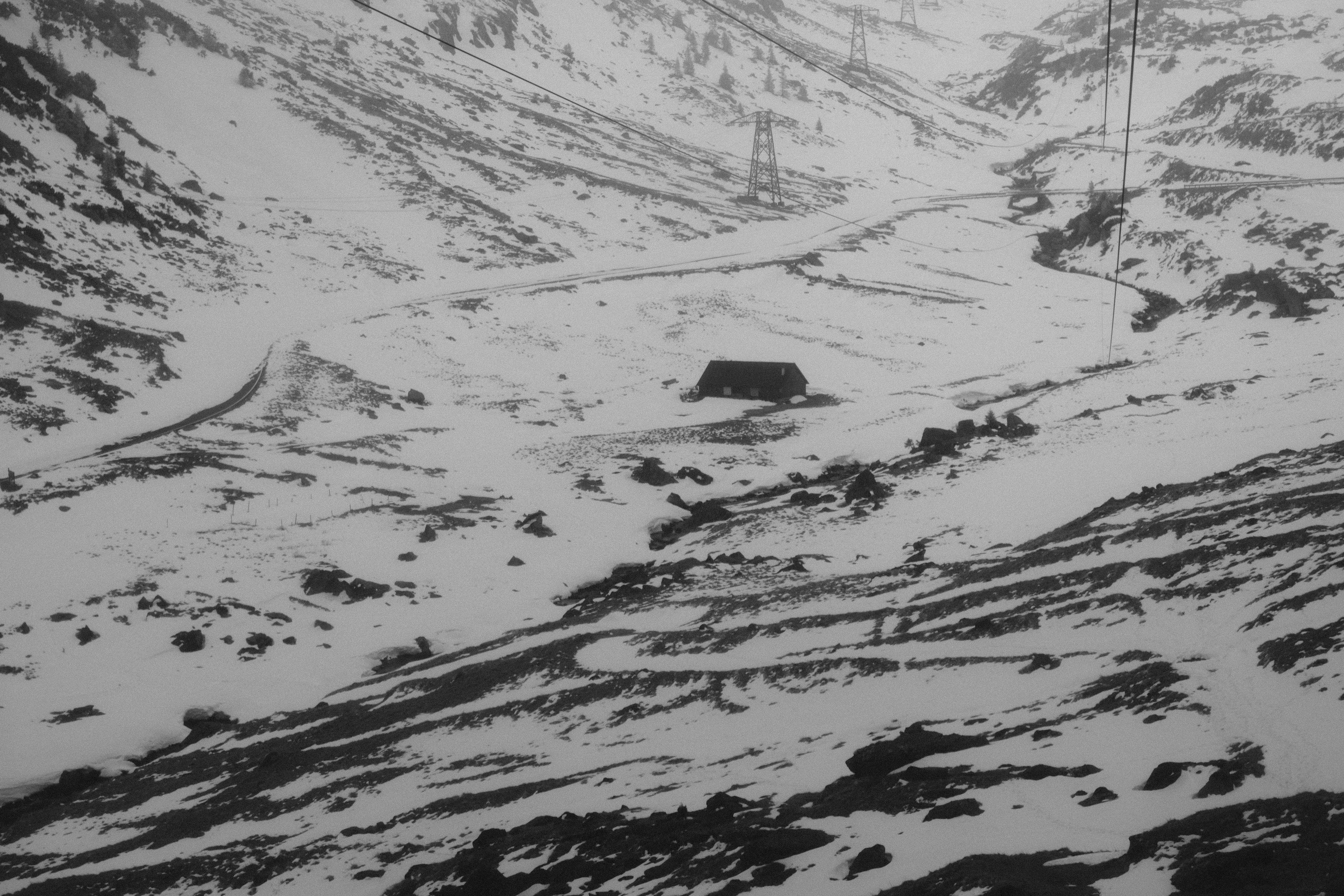 A lone cabin rests in a vast snowy landscape, surrounded by rugged terrain and distant power lines. The scene conveys a sense of isolation and tranquility.