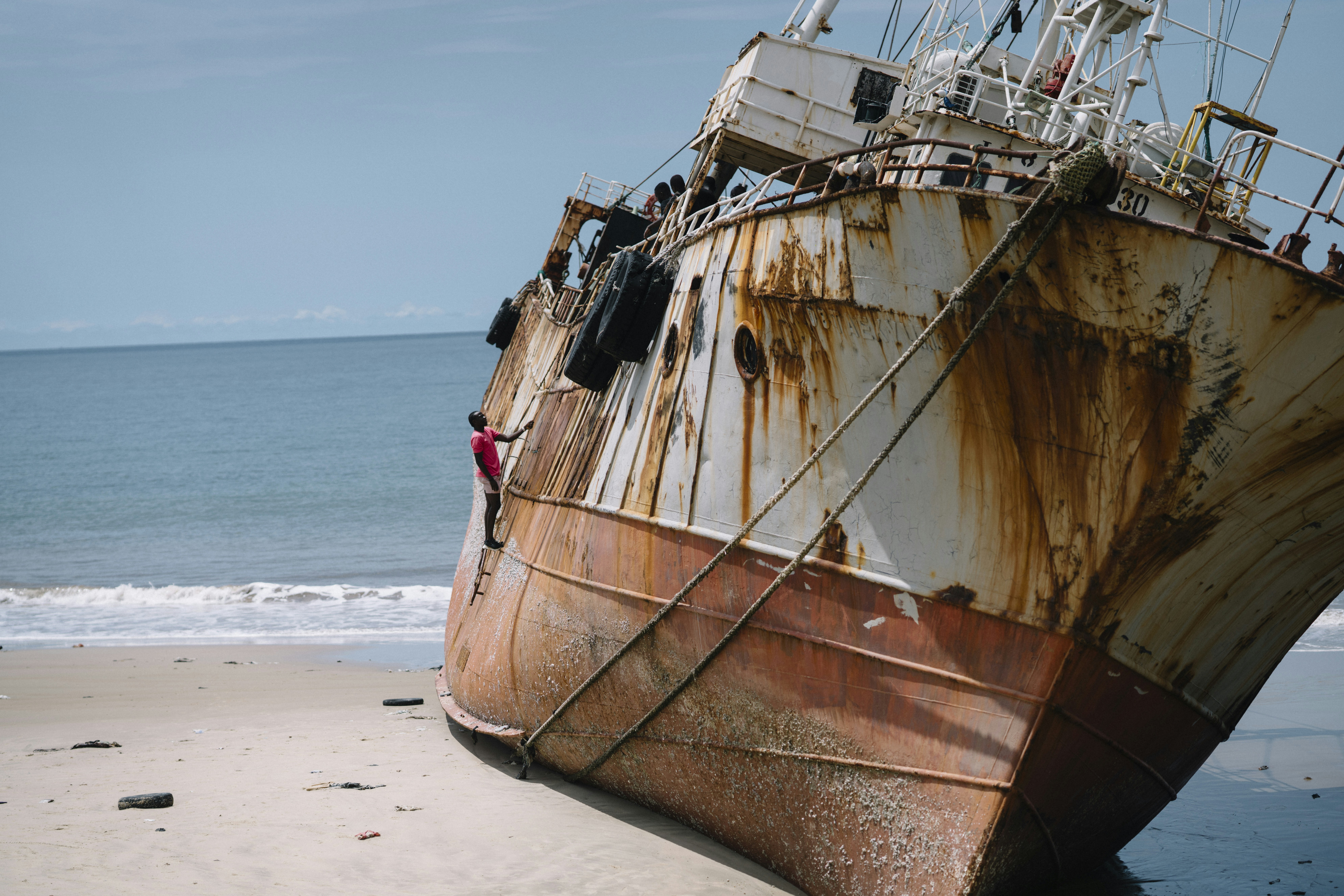 A climber scales the corroded hull of a beached ship, surrounded by serene waters and a clear sky.