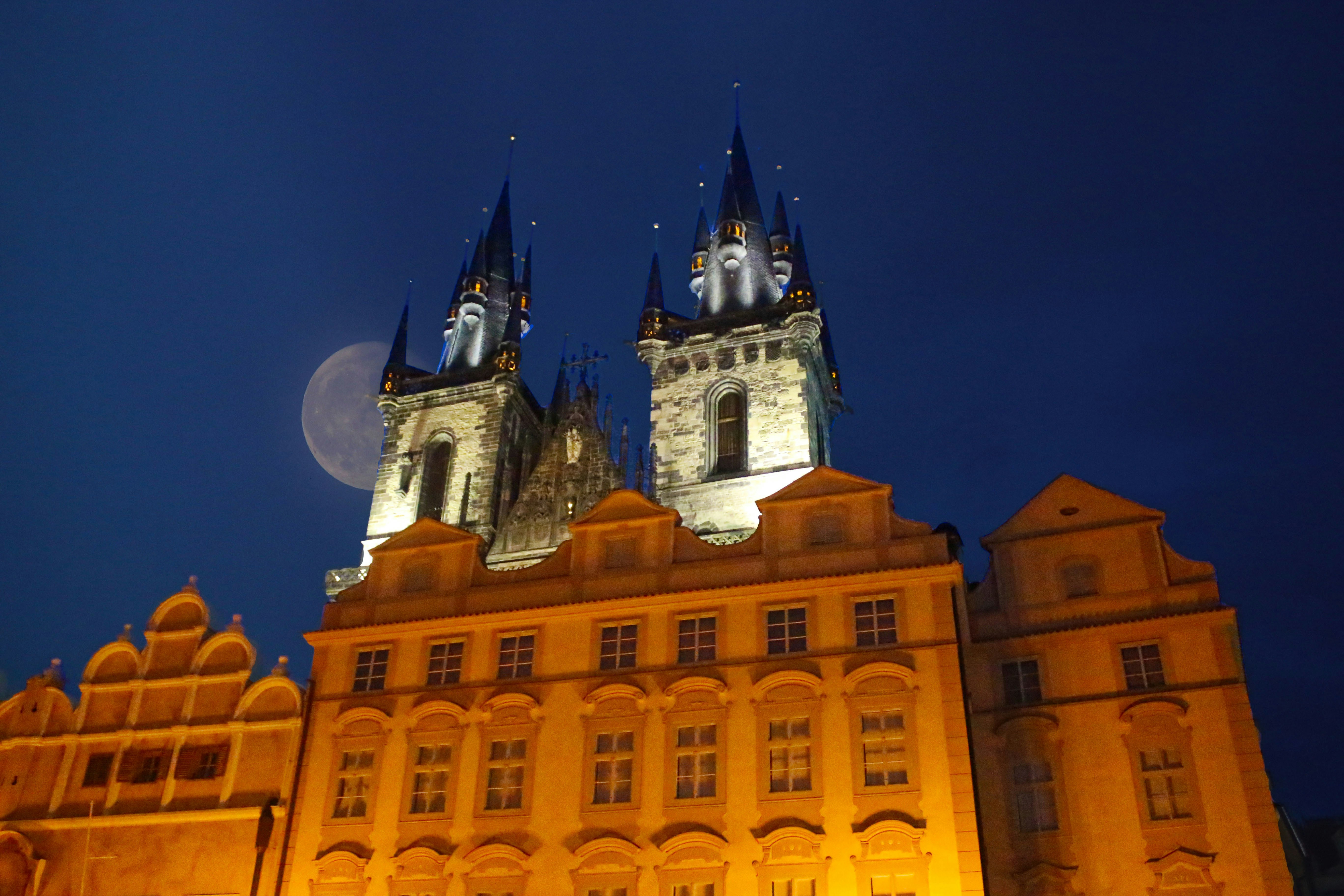 The Church of Our Lady before Týn glows under artificial lighting as an almost full moon rises behind one of its Gothic towers. This night scene captures the magical and mysterious atmosphere of Prague, where medieval history and natural beauty blend in perfect harmony. | Ornate buildings under a dark blue night sky