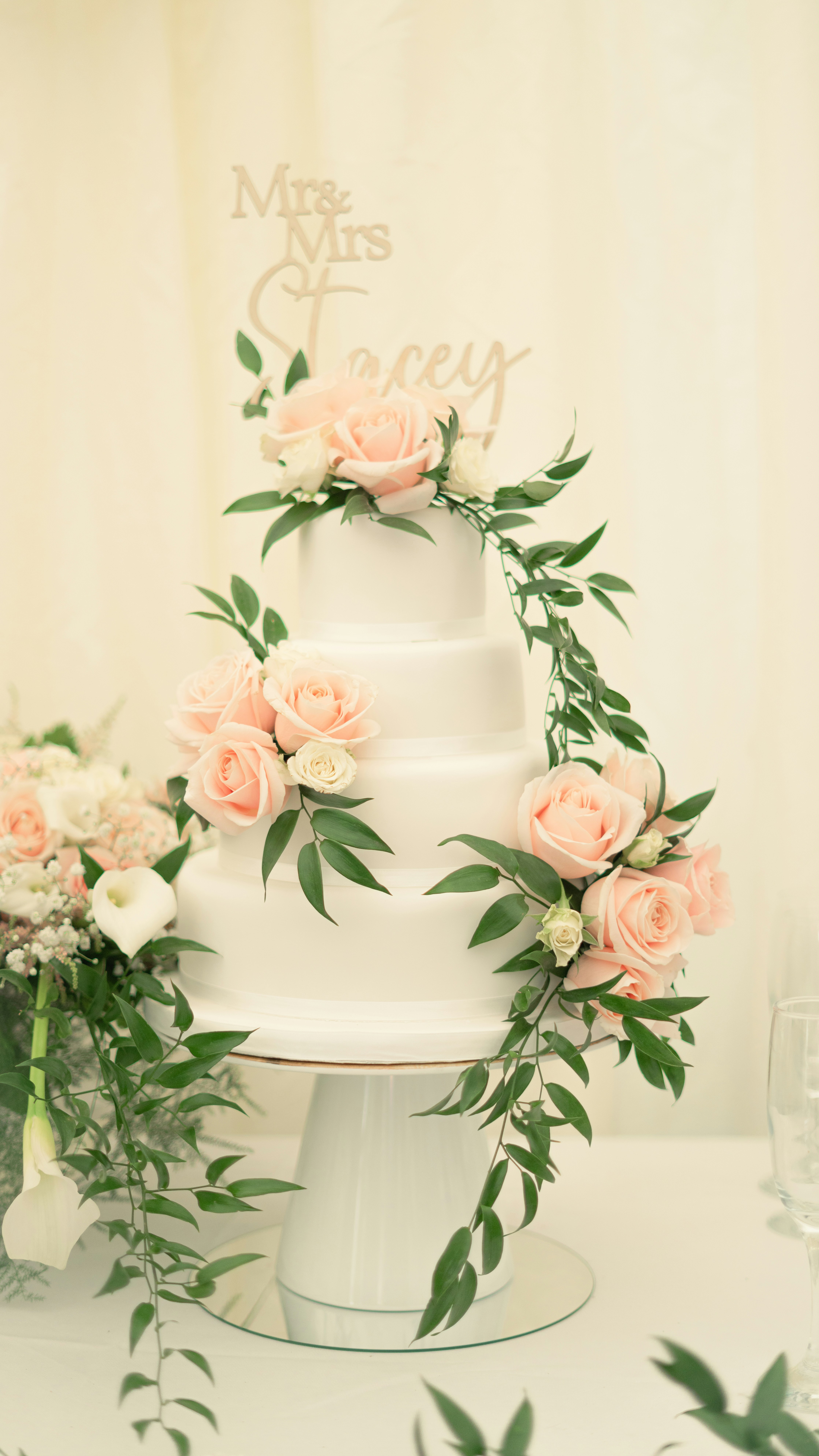 Three-tiered wedding cake adorned with delicate pink roses and greenery, topped with a custom wooden sign. Elegant floral arrangement complements the cake's design.