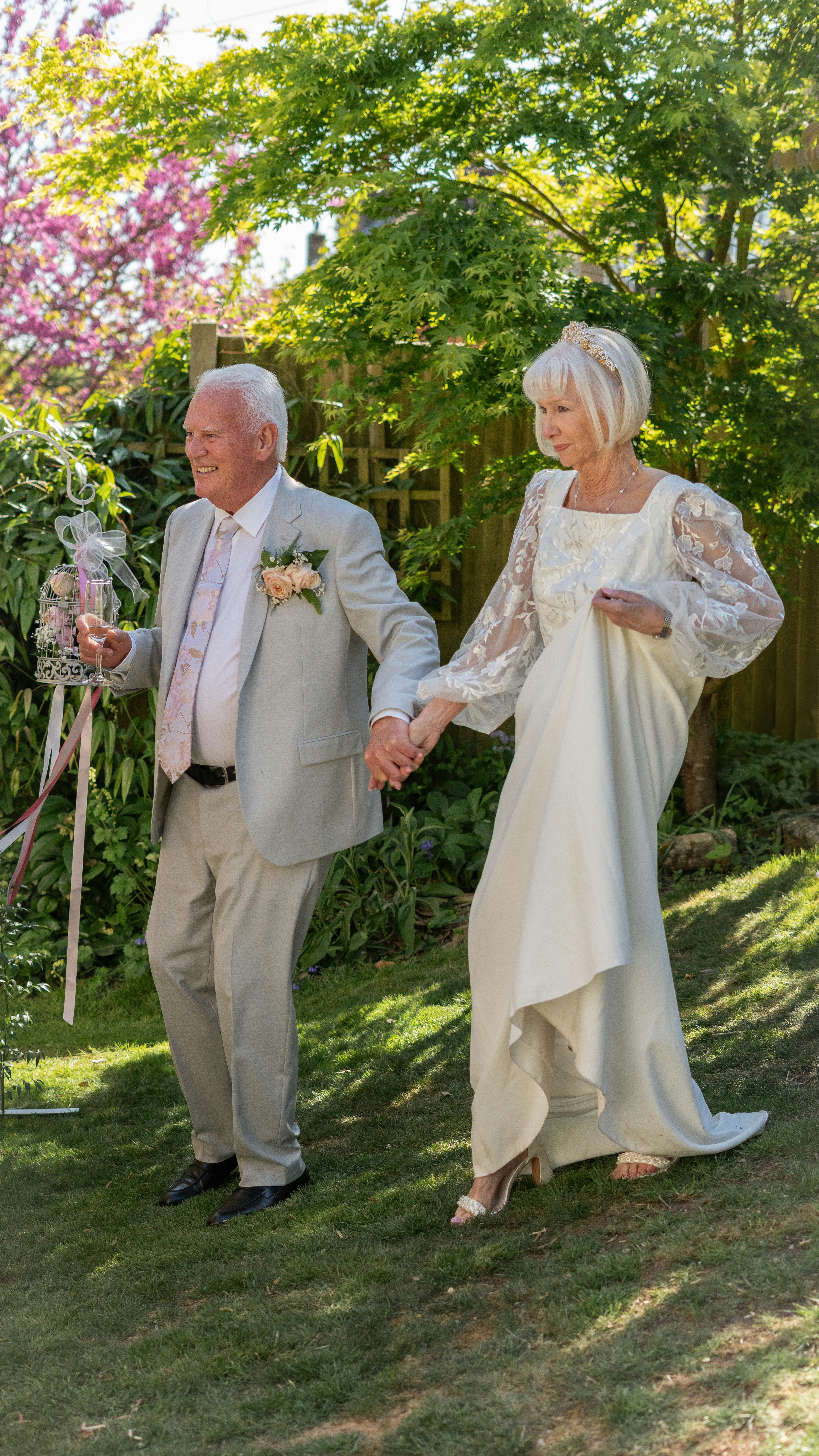 Elderly couple walking hand-in-hand in a garden, surrounded by vibrant greenery and blooming flowers.