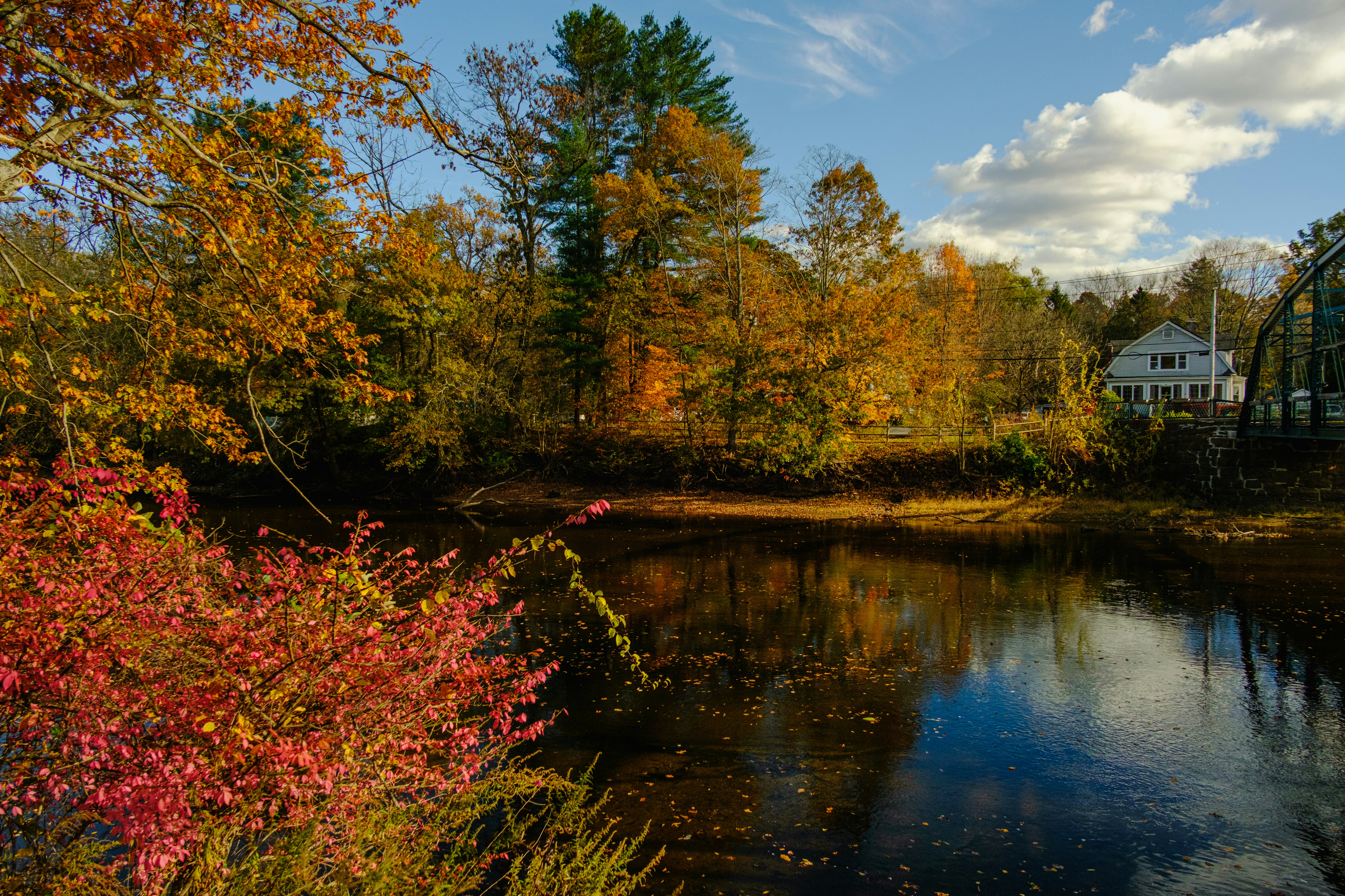 Vibrant autumn foliage frames a tranquil river reflecting a clear blue sky and scattered clouds. A charming house peeks through the trees.