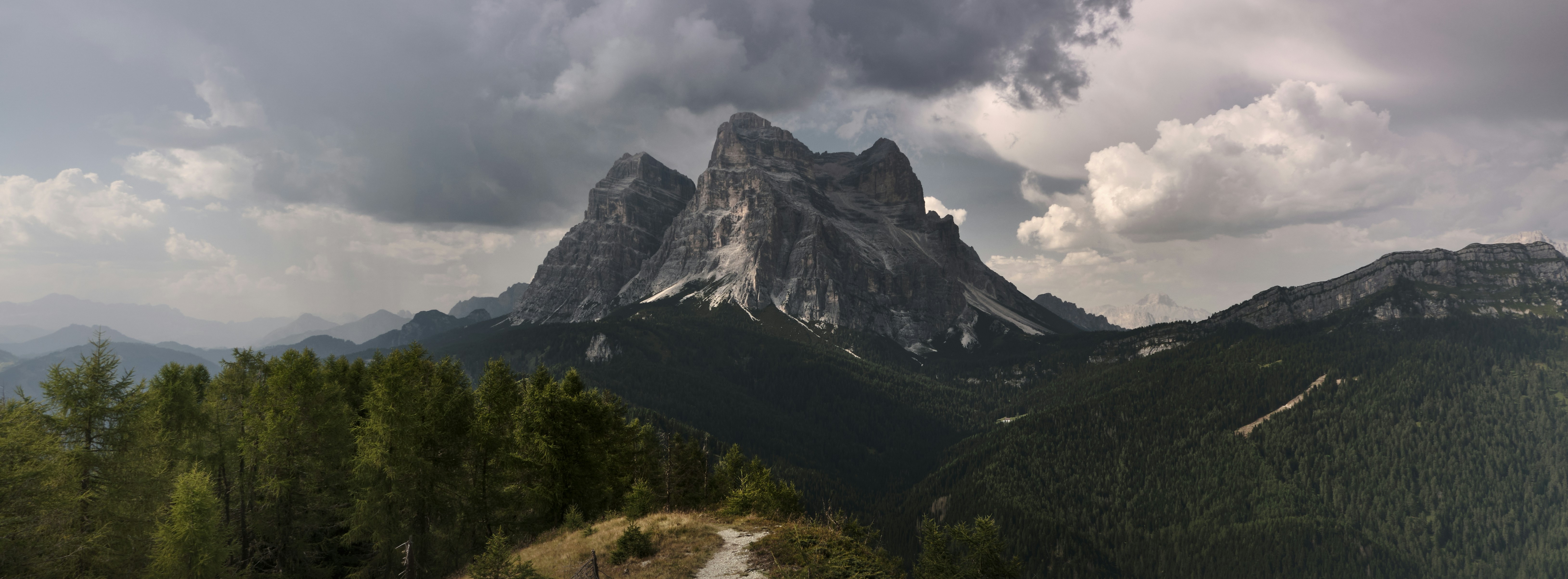 Dramatic mountain range under a moody sky, showcasing rugged peaks and lush greenery in the foreground.