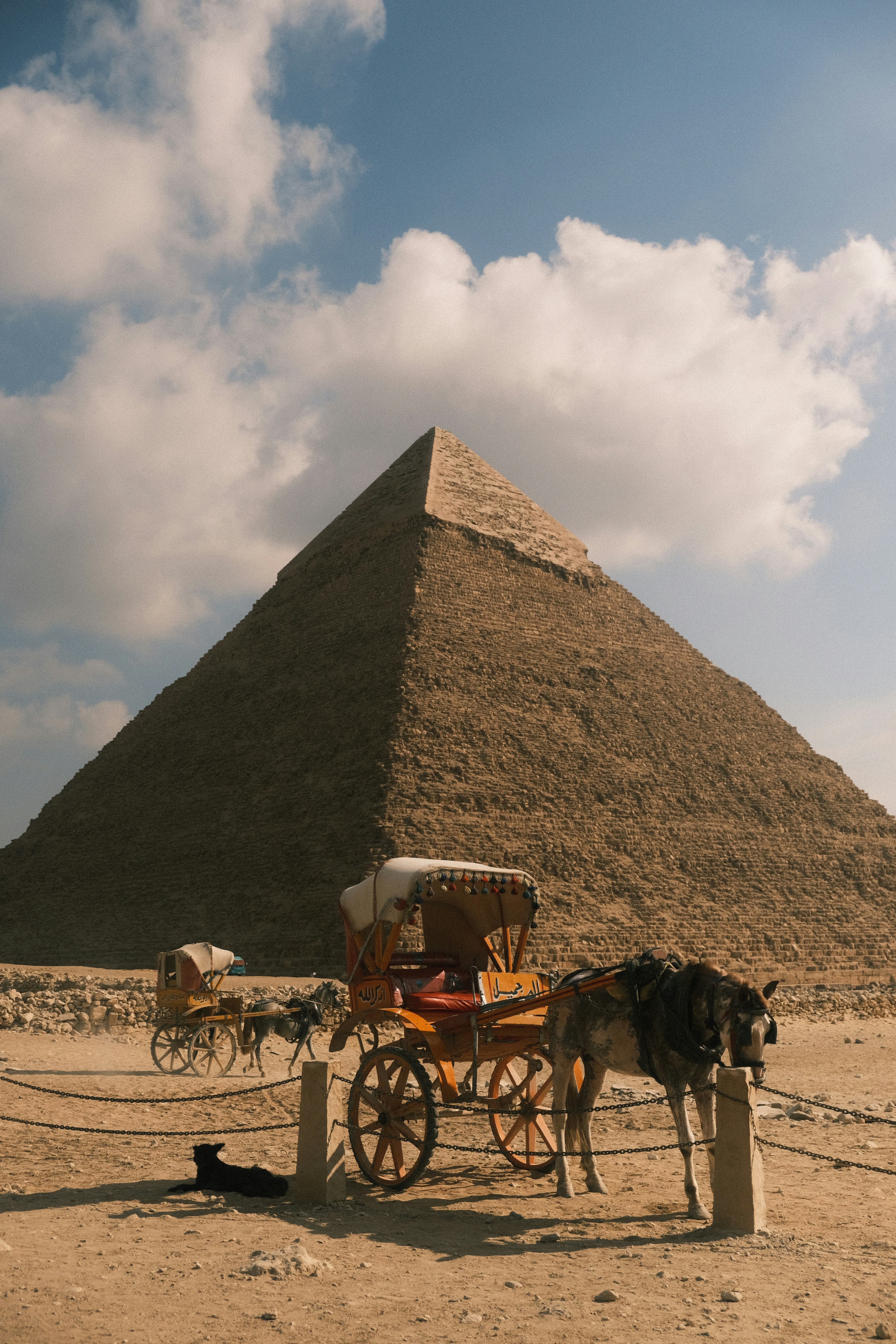 A horse-drawn cart rests in the foreground with a pyramid looming majestically in the background, under a partly cloudy sky.