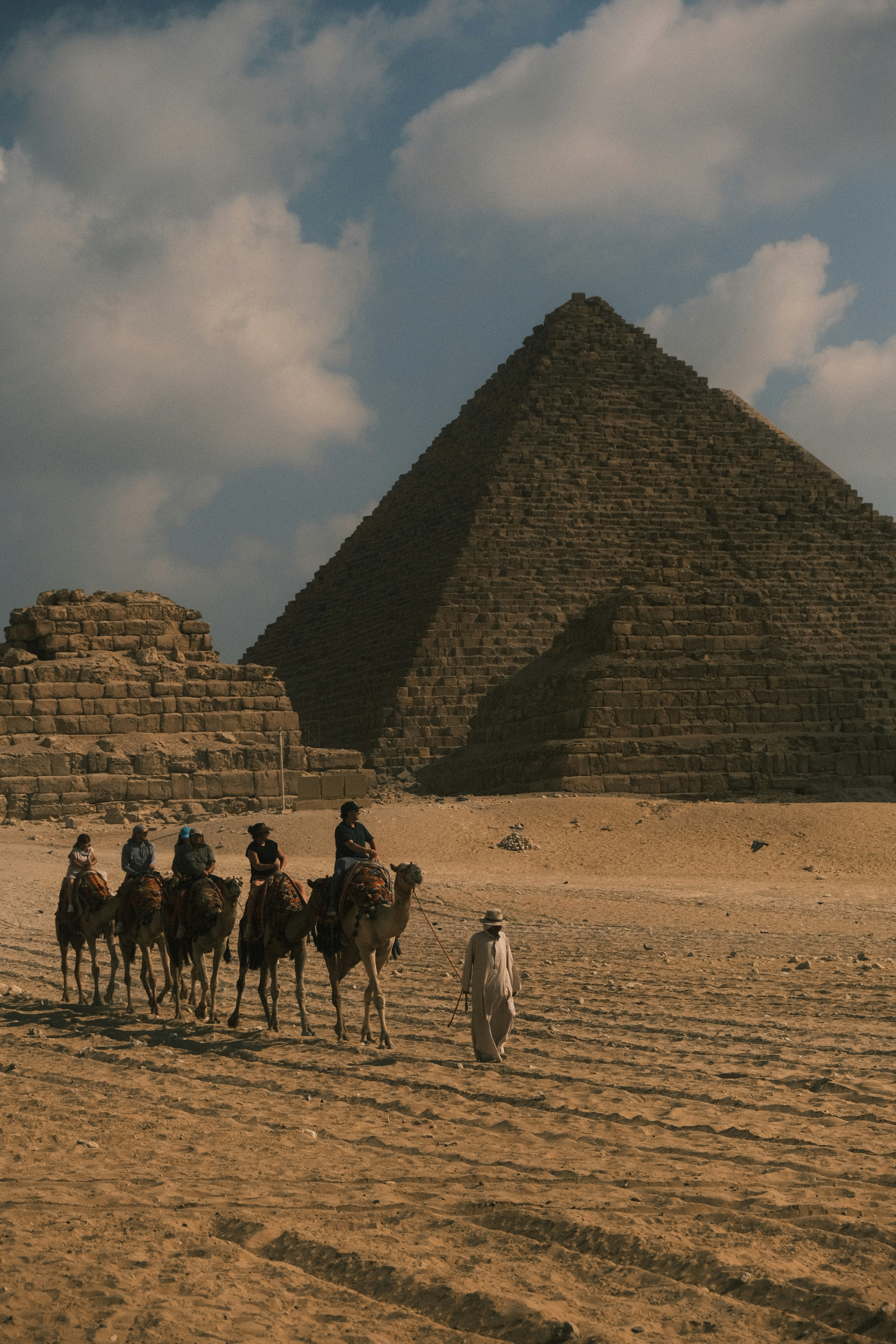 A caravan of camels traversing the sandy expanse near the Great Pyramid, with a figure walking ahead. The scene captures the timeless connection between humanity and ancient architecture.