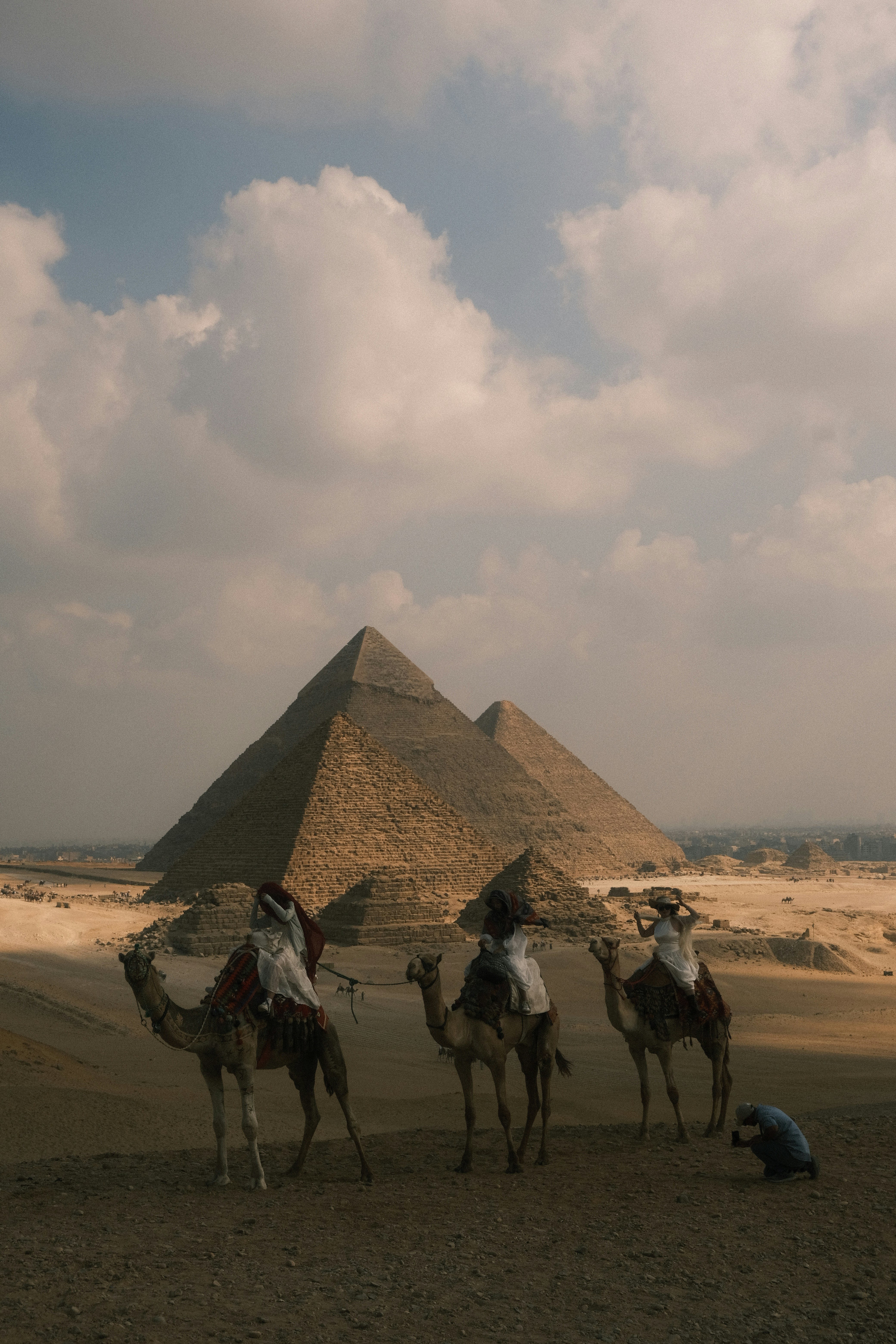 Three camels with riders stand before the Great Pyramids, set against a dramatic sky filled with clouds. A figure kneels in the foreground, adding depth to the scene.