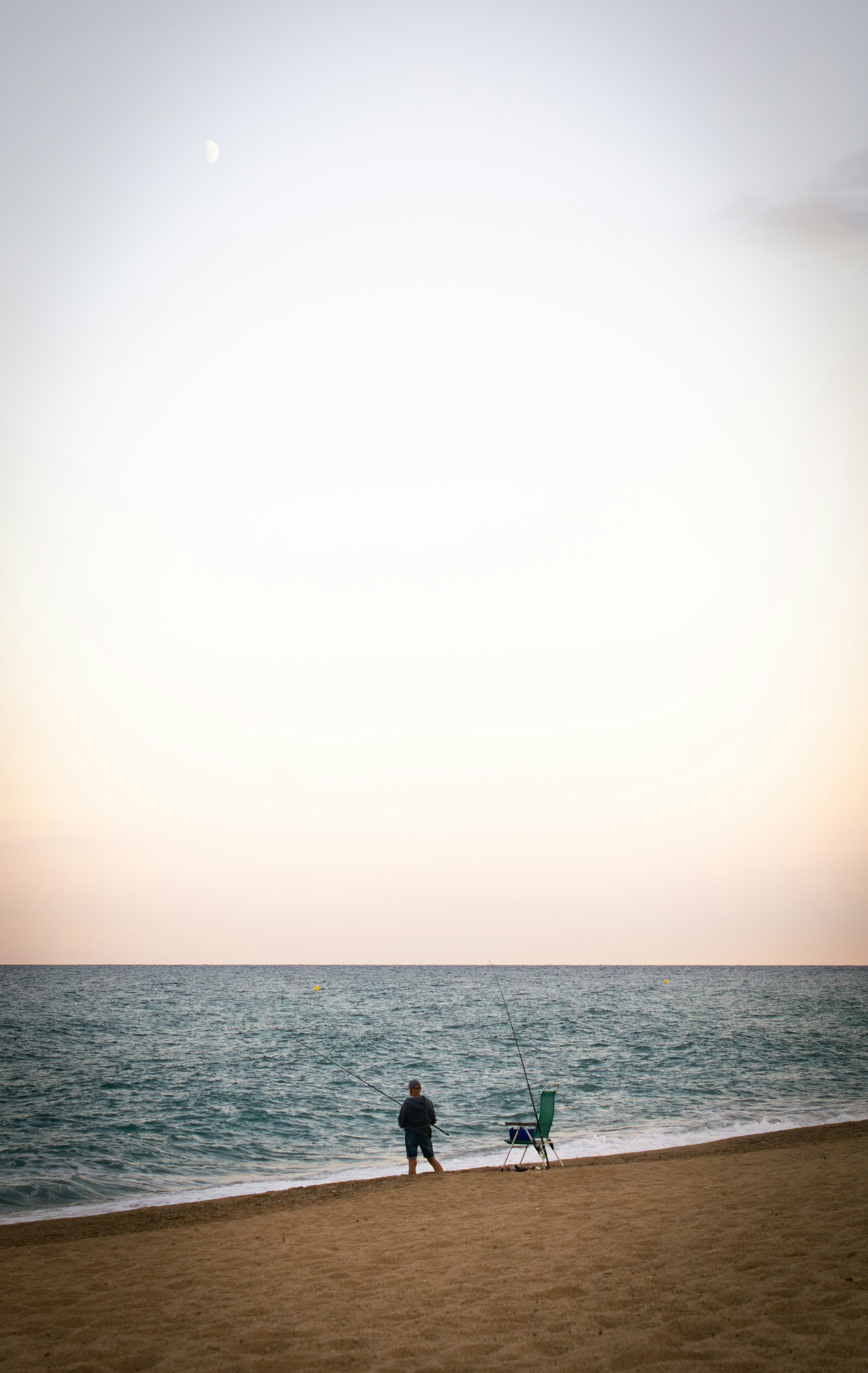 A person fishing on a quiet beach. A fishing rod and chair sit on the sand next to the calm ocean under a soft sky with a crescent moon. | Man standing on beach next to green chair
