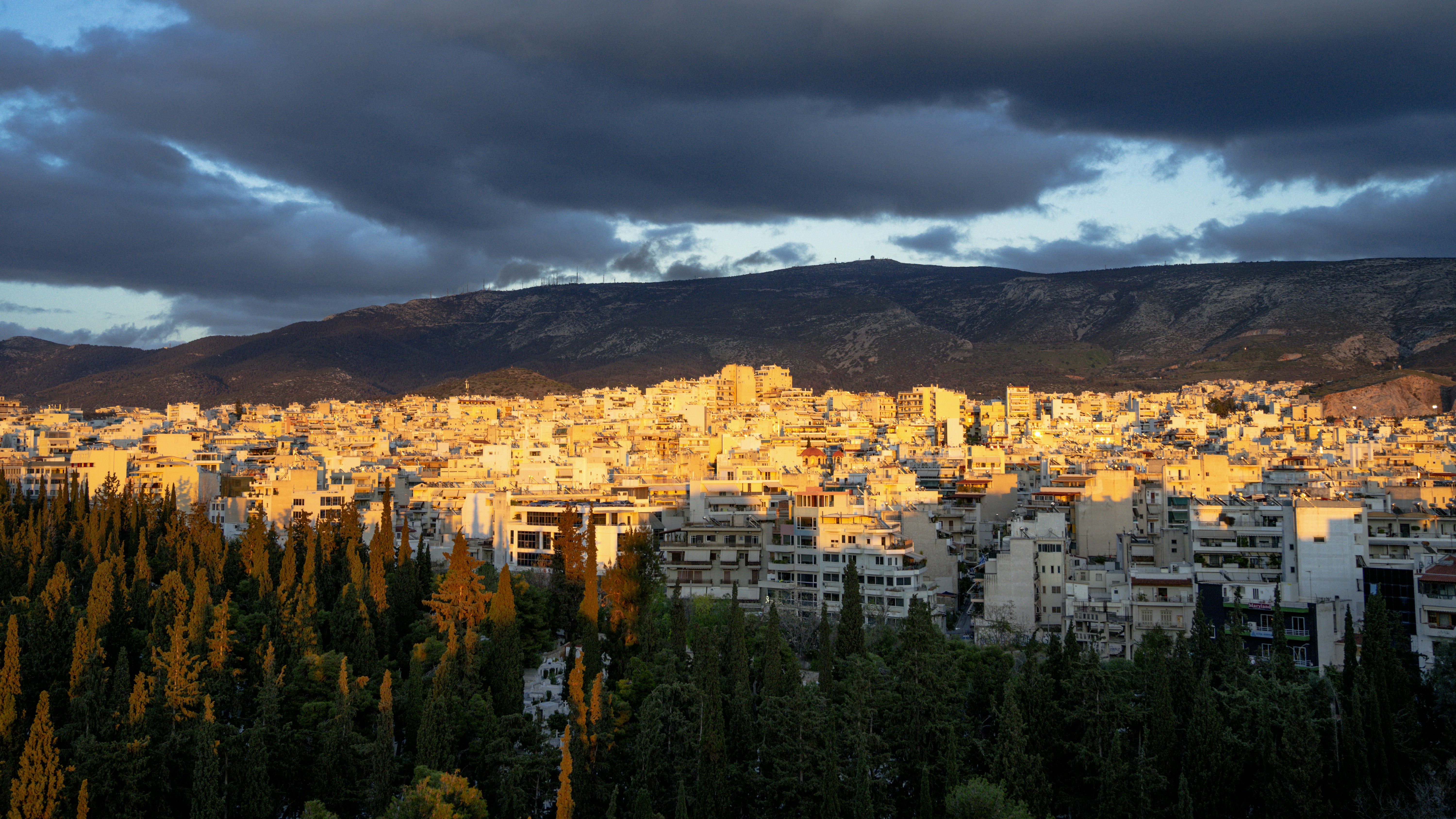 A panoramic view of a cityscape bathed in warm golden light, contrasting with the surrounding lush greenery and dramatic mountain backdrop.