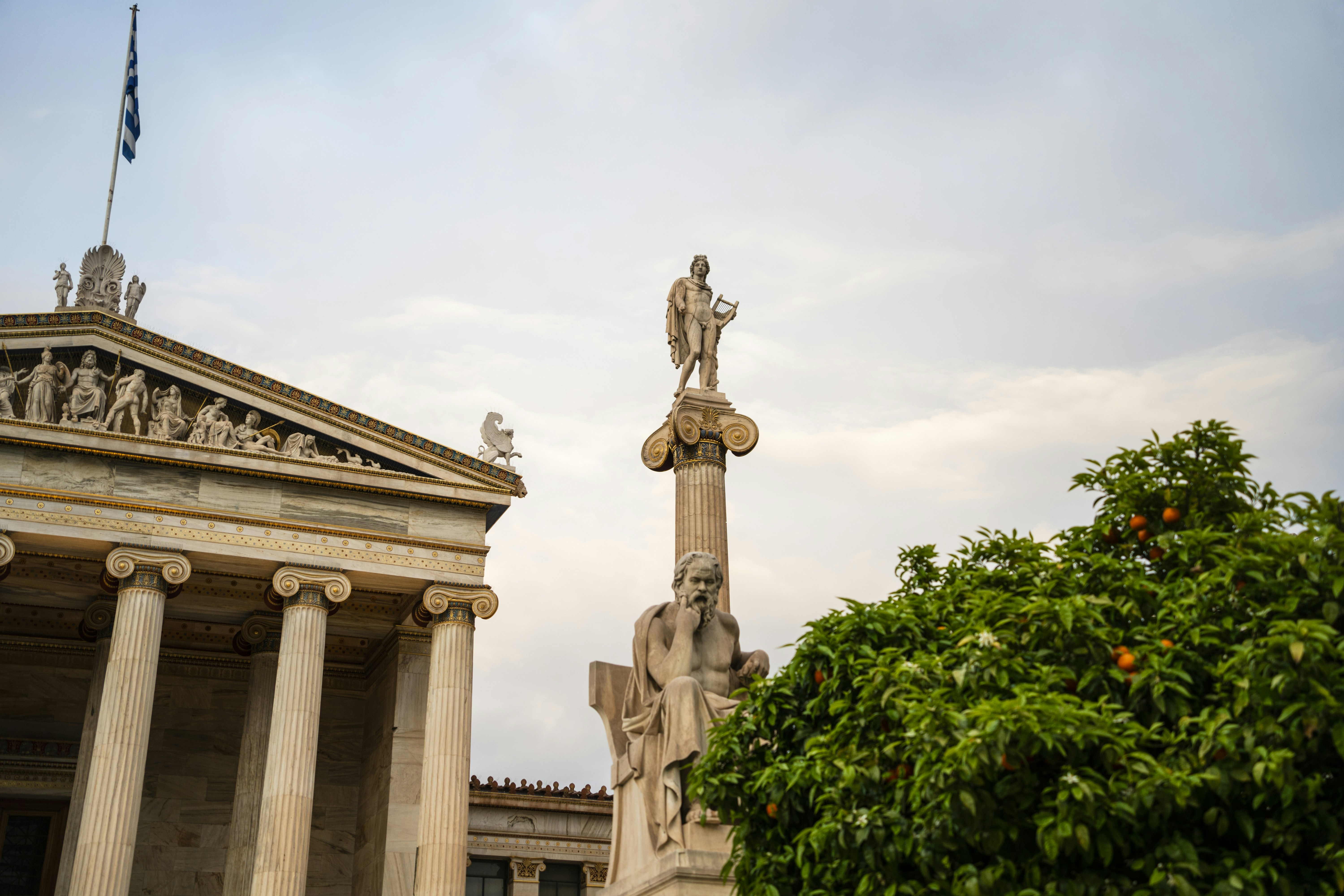 A neoclassical building adorned with statues, featuring a prominent figure atop a column alongside a lush orange tree, symbolizing the blend of culture and nature.