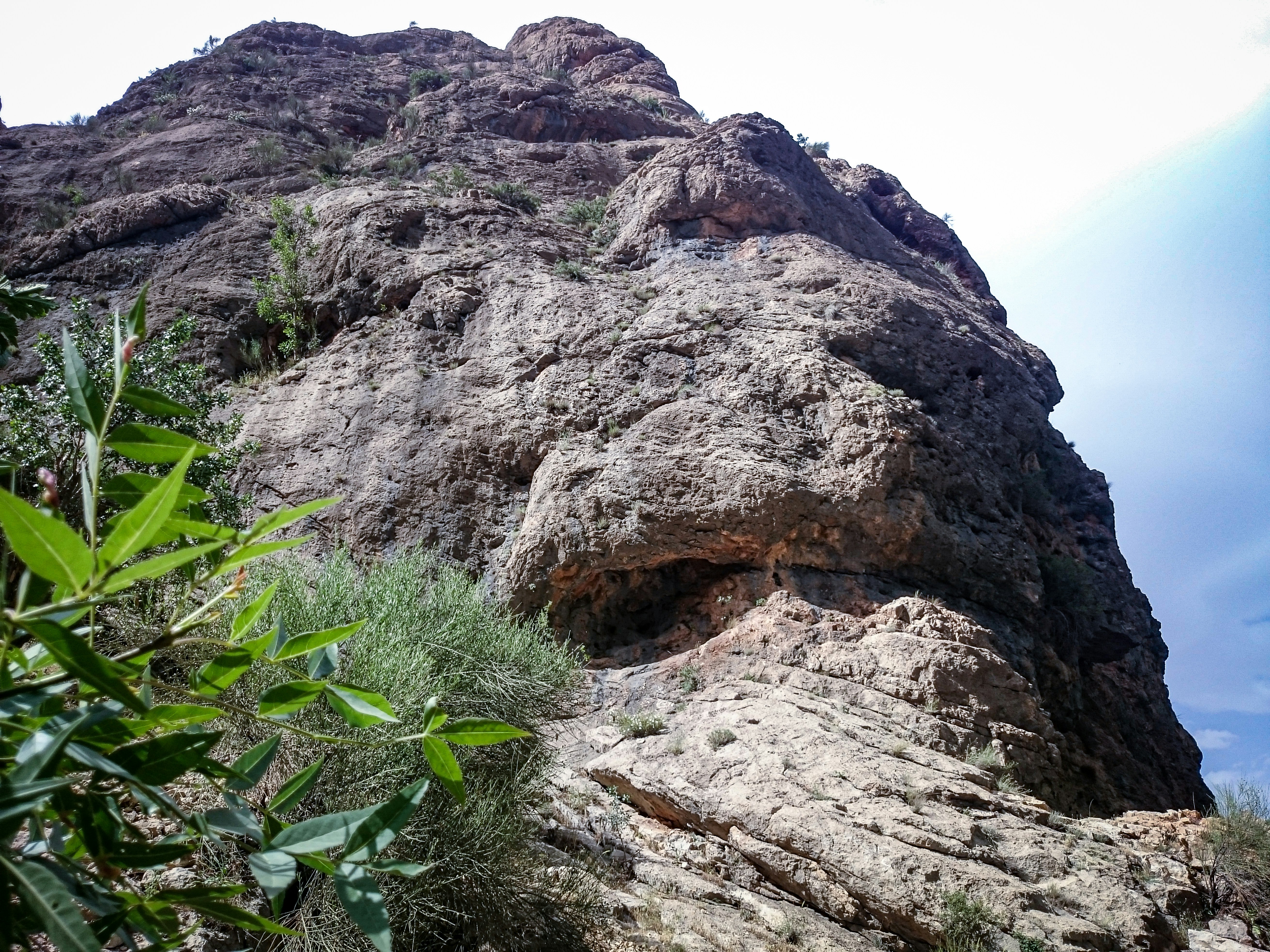 Rugged Cliffs — Lost Paradise — May 15, 2015 (#0018) This photograph, taken from a low perspective, emphasizes the towering height and textured, layered geology of a large cliffside. The harsh sunlight highlights the cracks and crevices of the brownish-grey rock. In the bottom-left corner, vibrant green bushes provide a soft contrast to the hard rock, framing the scene. A small patch of pale sky is visible in the upper right. Sony Xperia Z1 (C6903) • 4.9 mm • f/2.0 • 1/1250 s • ISO 50 Full-res download: mofaeye.com/18 (link in bio) #BeheshteGomshodeh #LostParadise #RockFormation #Cliffs | A large rock formation with green foliage