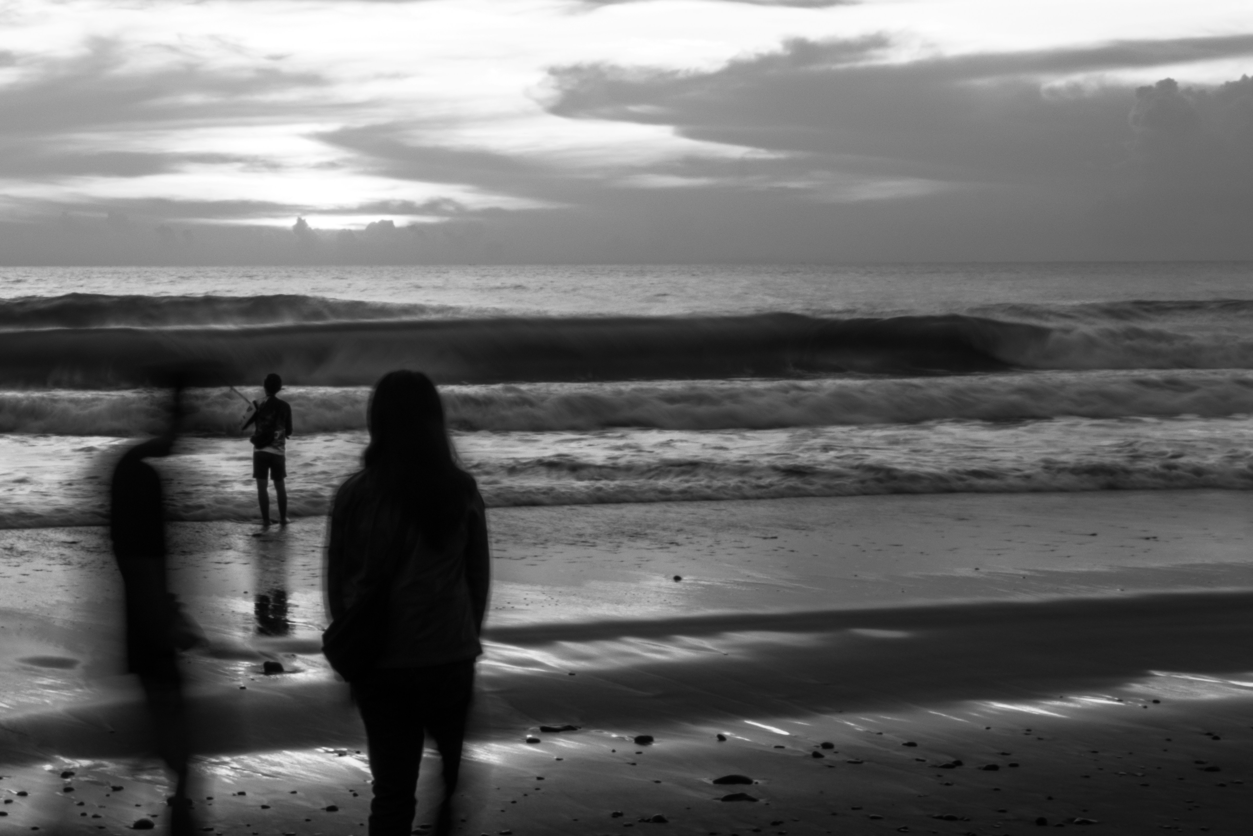 Silhouetted figures stand on a beach as waves crash under a moody sky, creating a dynamic interplay of movement and stillness.