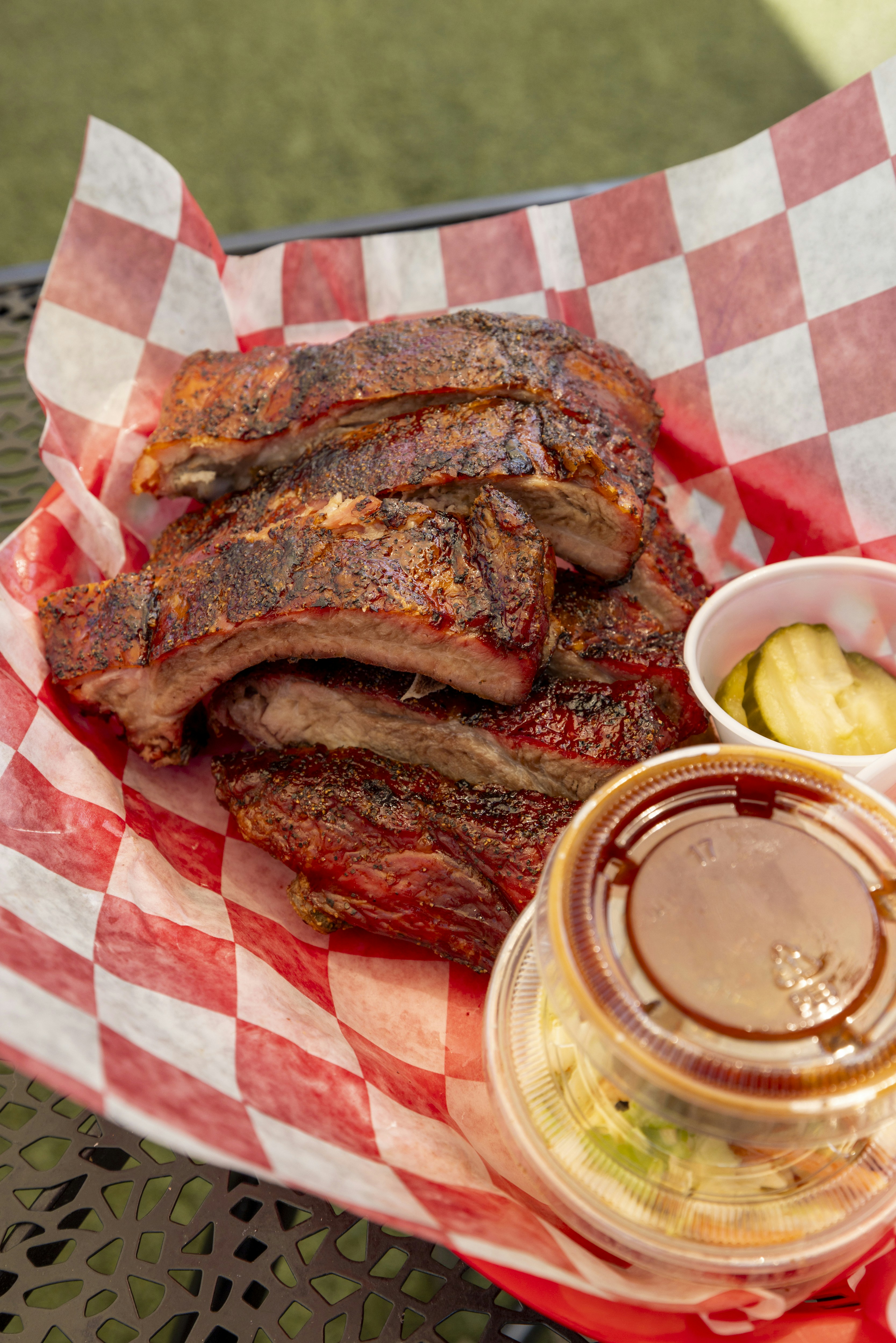Three succulent barbecue ribs resting on a checkered paper, accompanied by pickles and sauce in containers.