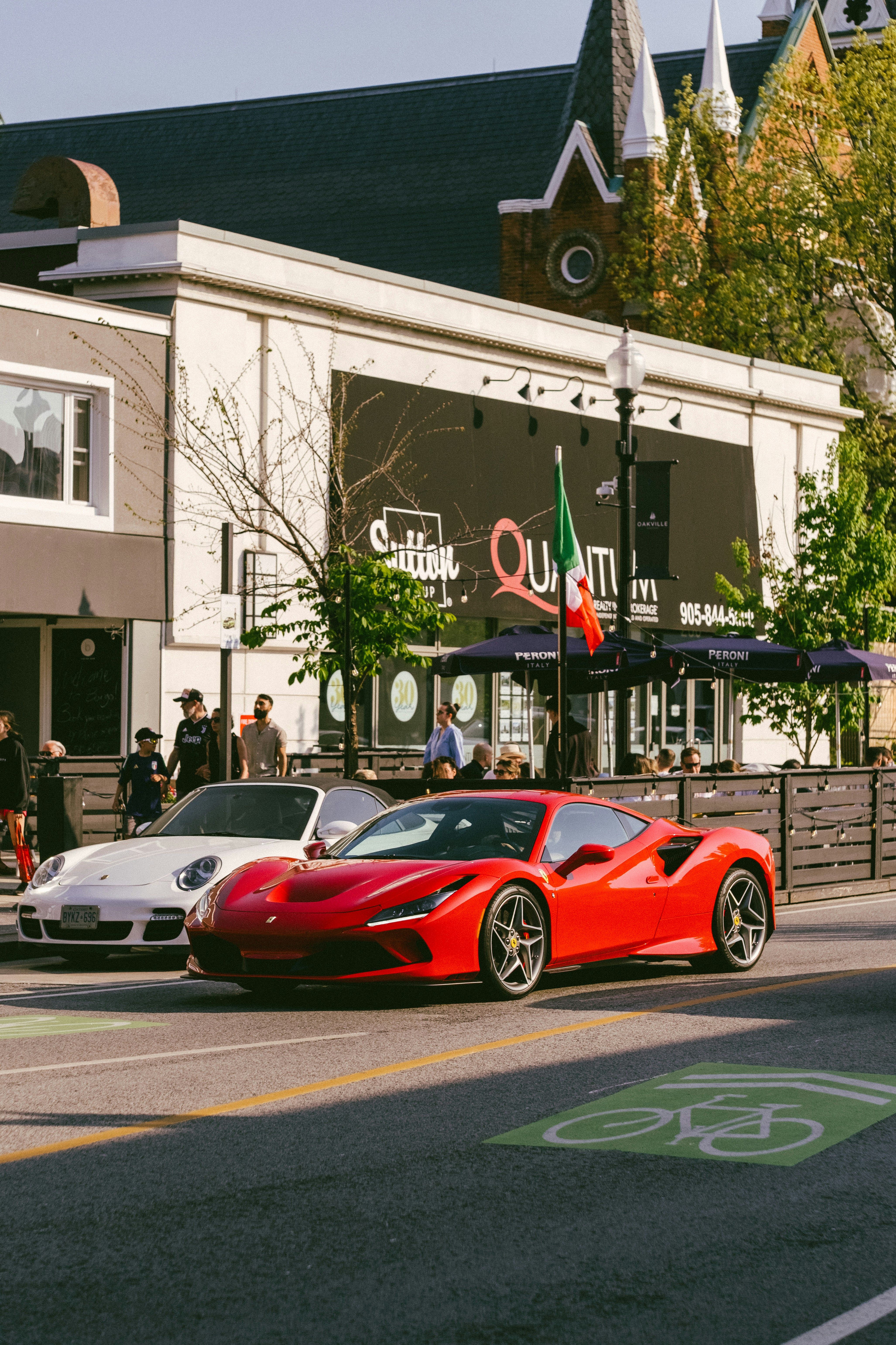 A vibrant red sports car and a sleek white coupe parked on a lively city street, surrounded by greenery and urban architecture.