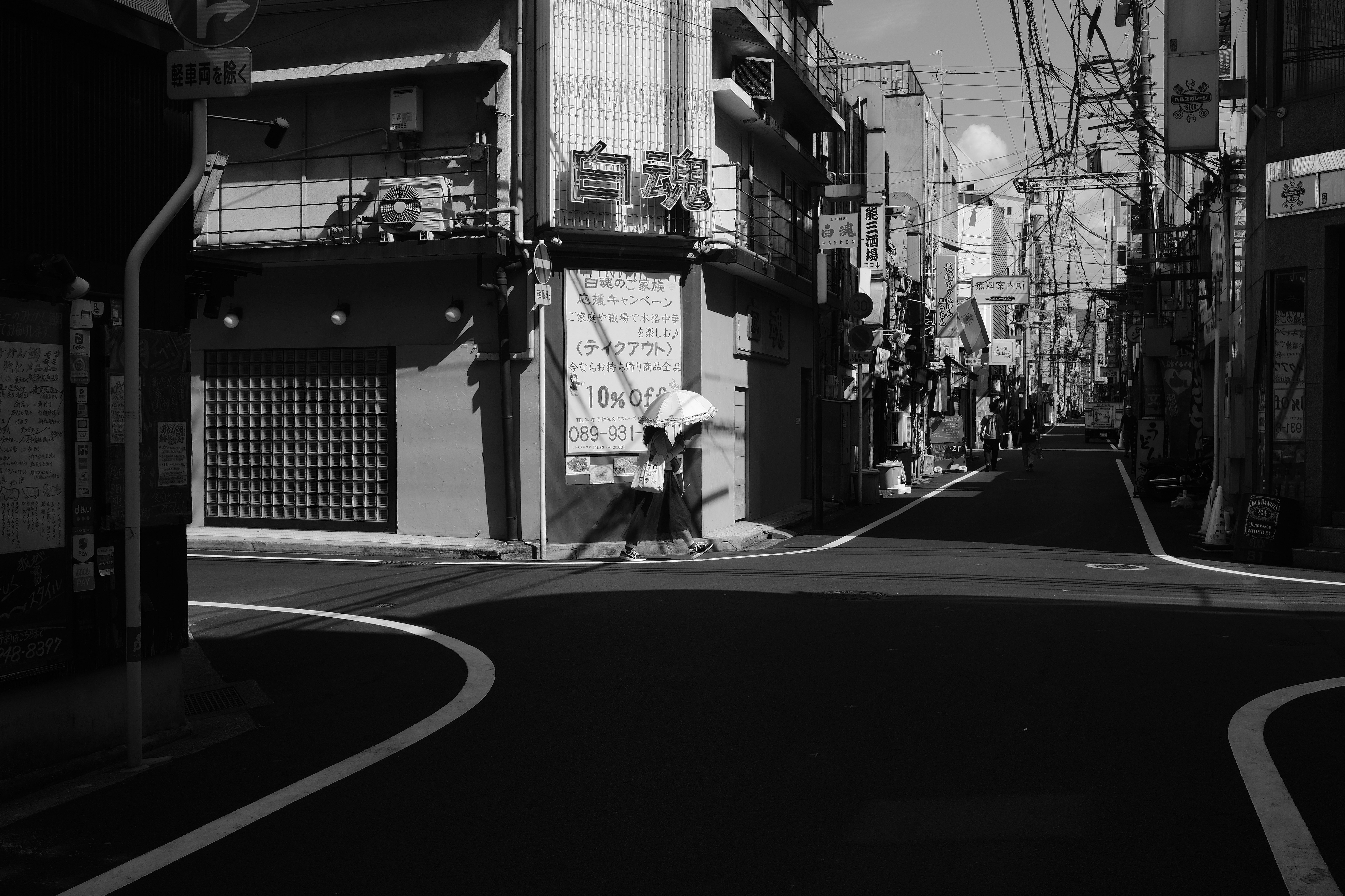 A solitary figure with an umbrella walks through a quiet city street, flanked by buildings and power lines in a monochromatic setting.