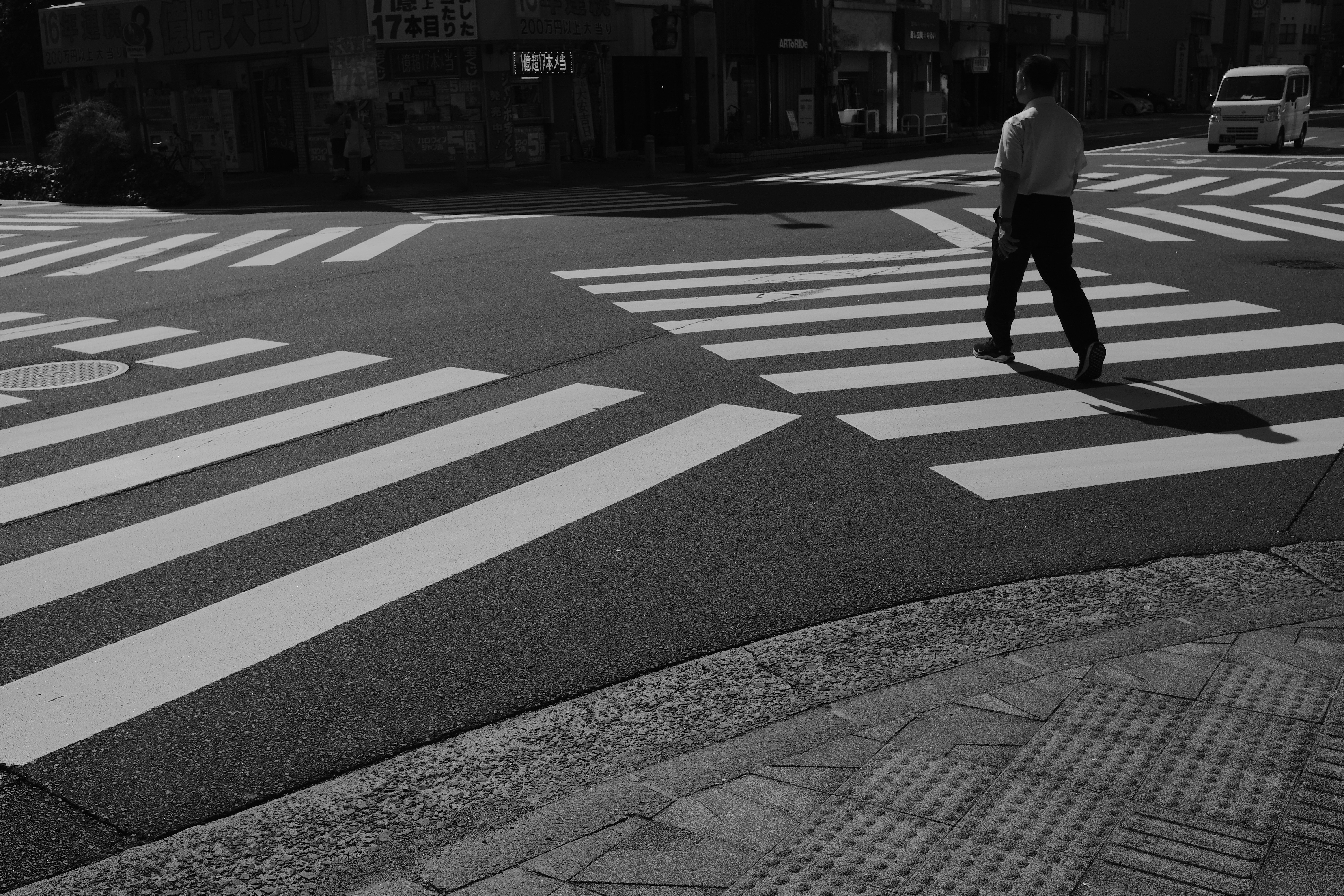 A lone pedestrian crosses a starkly patterned crosswalk in an urban setting, emphasizing the interplay of light and shadow.