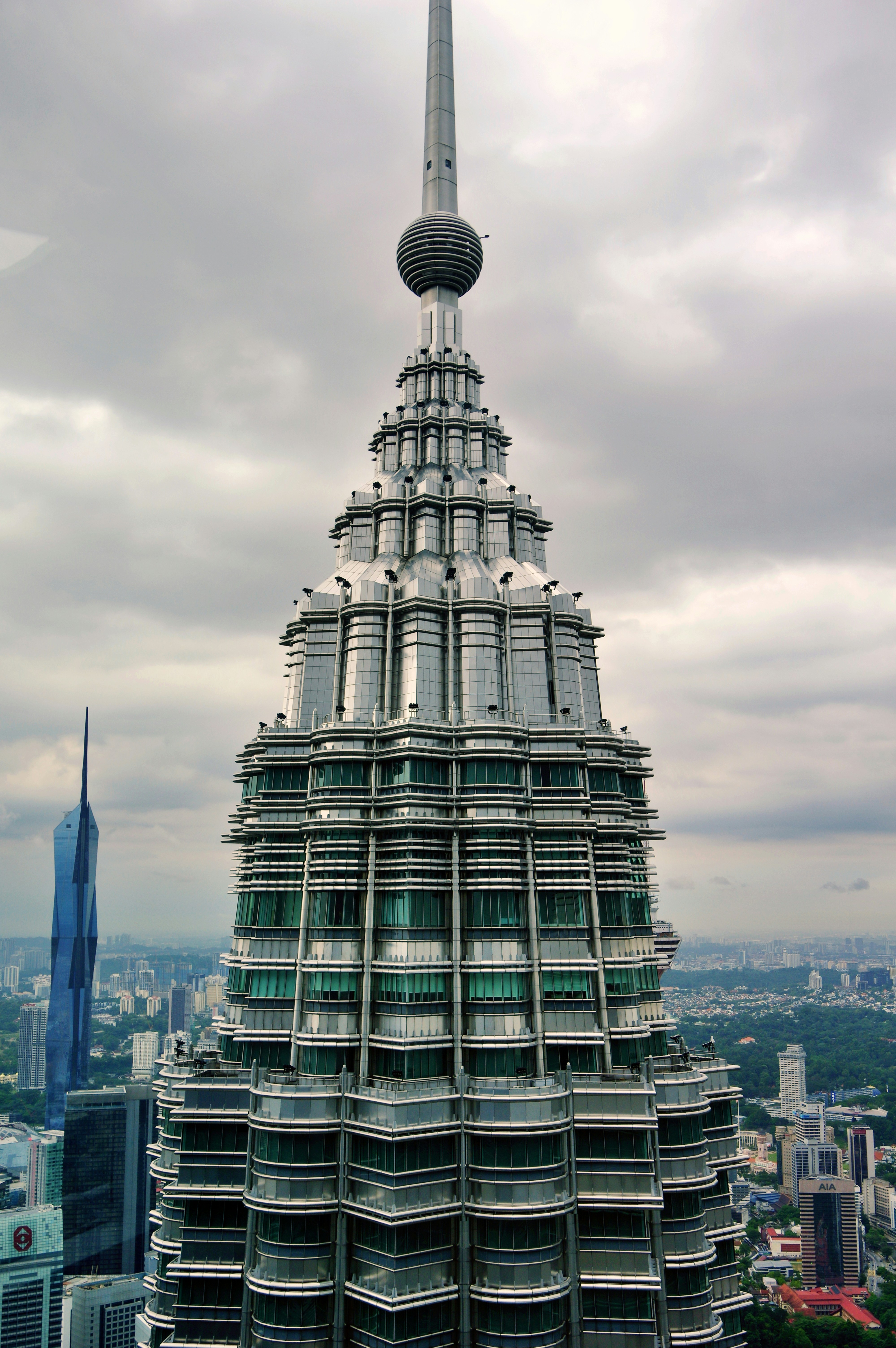 The intricate spire of the Petronas Towers rises dramatically against a cloudy sky, showcasing the marvel of modern engineering. The surrounding skyline adds depth to the urban landscape.